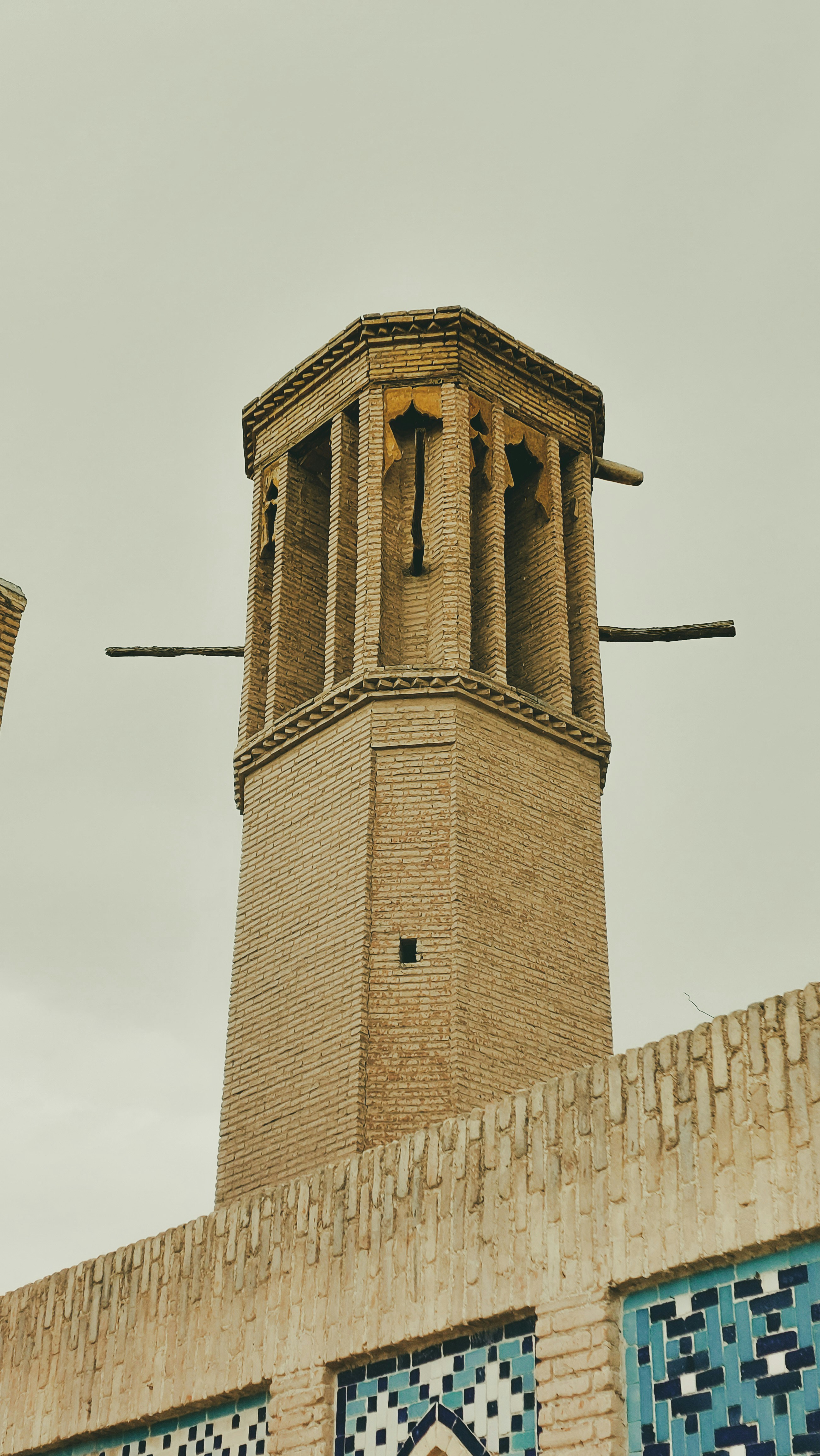 Beige brick wind tower rises above a low wall adorned with turquoise mosaic tiles, set against a pale sky.