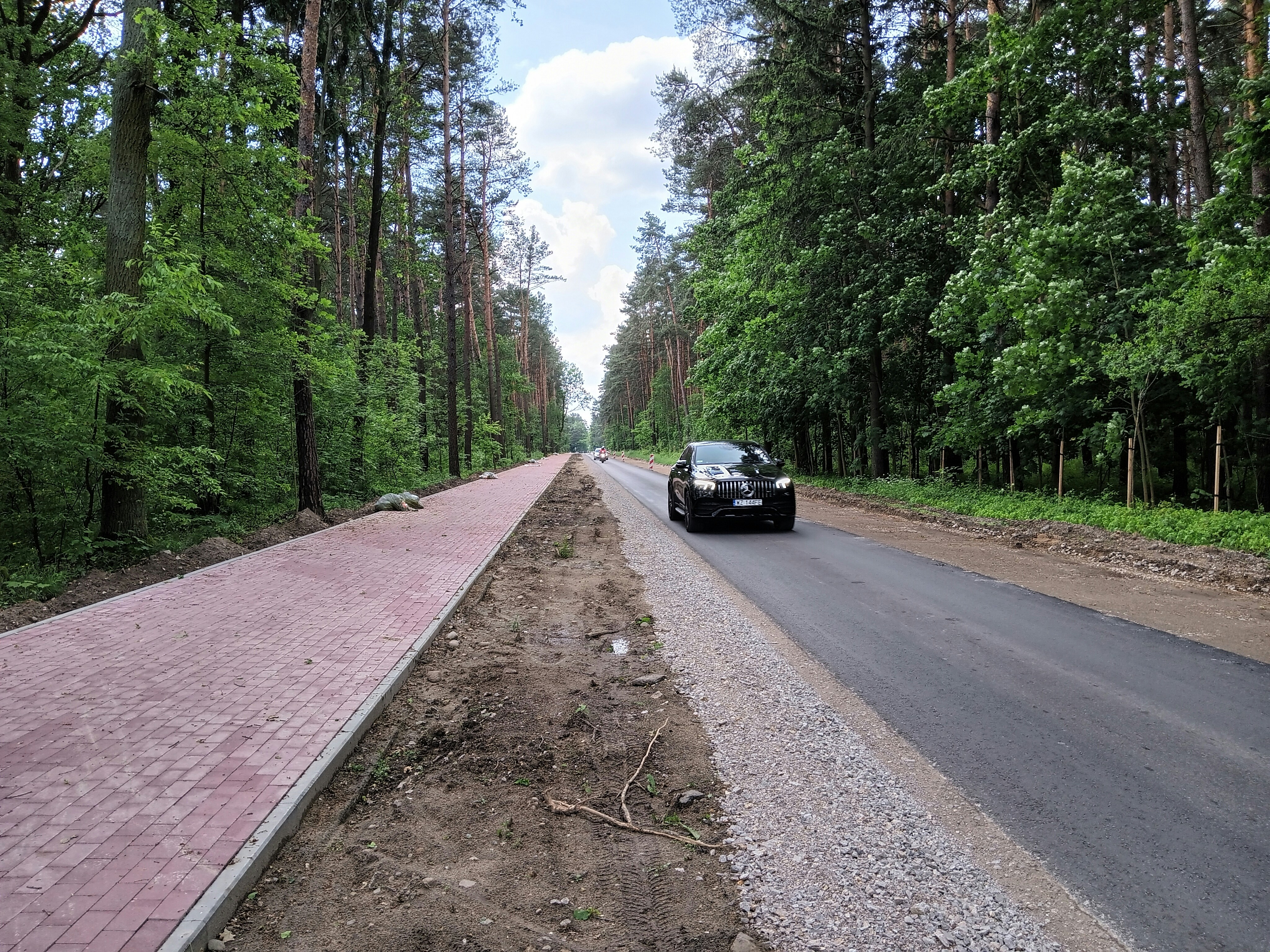 Black SUV drives along a freshly paved, tree-lined road with a pink brick sidewalk on the left. The scene captures ongoing road work and a calm rural corridor.