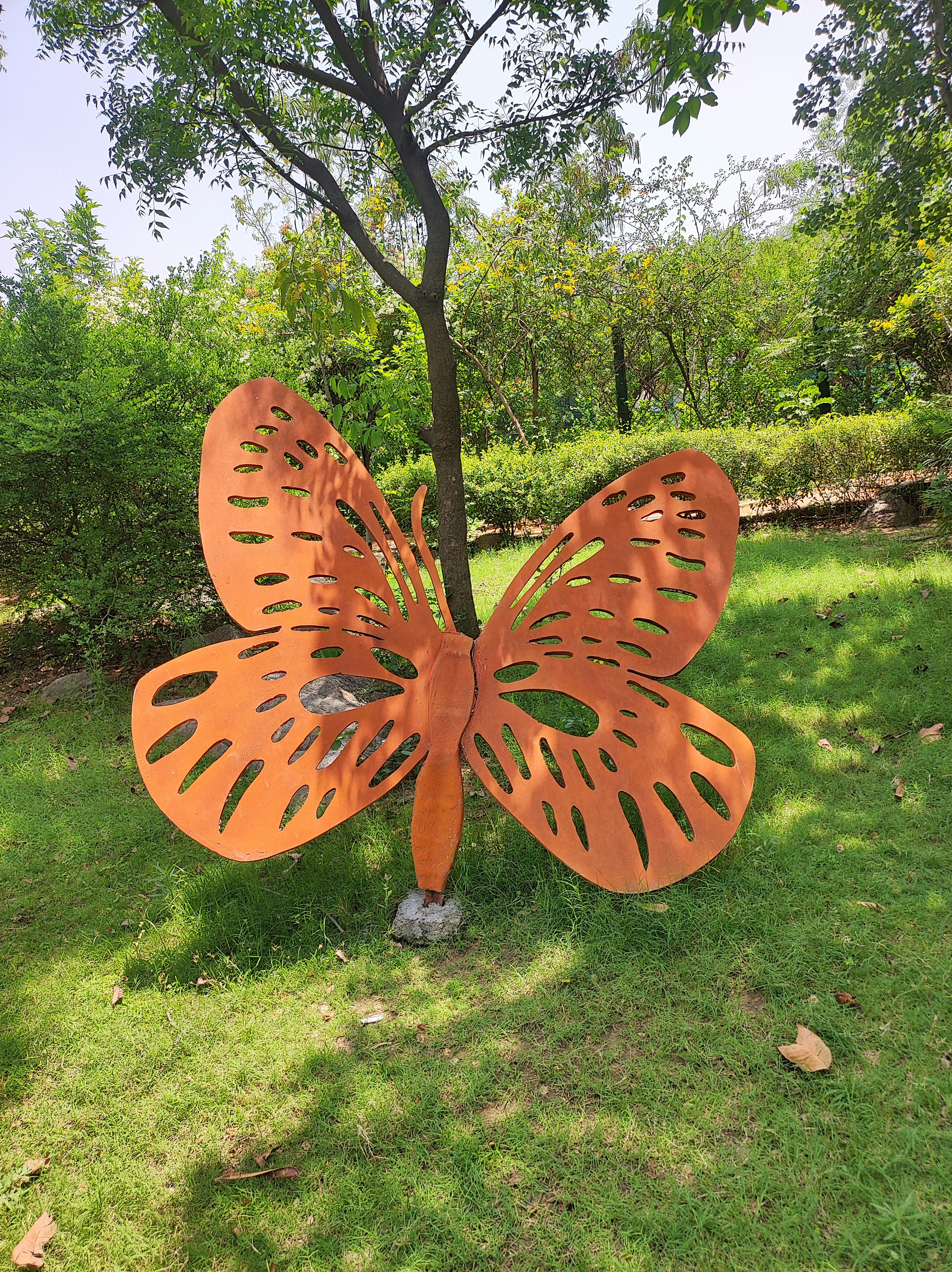 Copper butterfly sculpture with cut-out patterns sits on a small base in a sunlit park. The scene highlights an outdoor sculpture amid grass and trees.