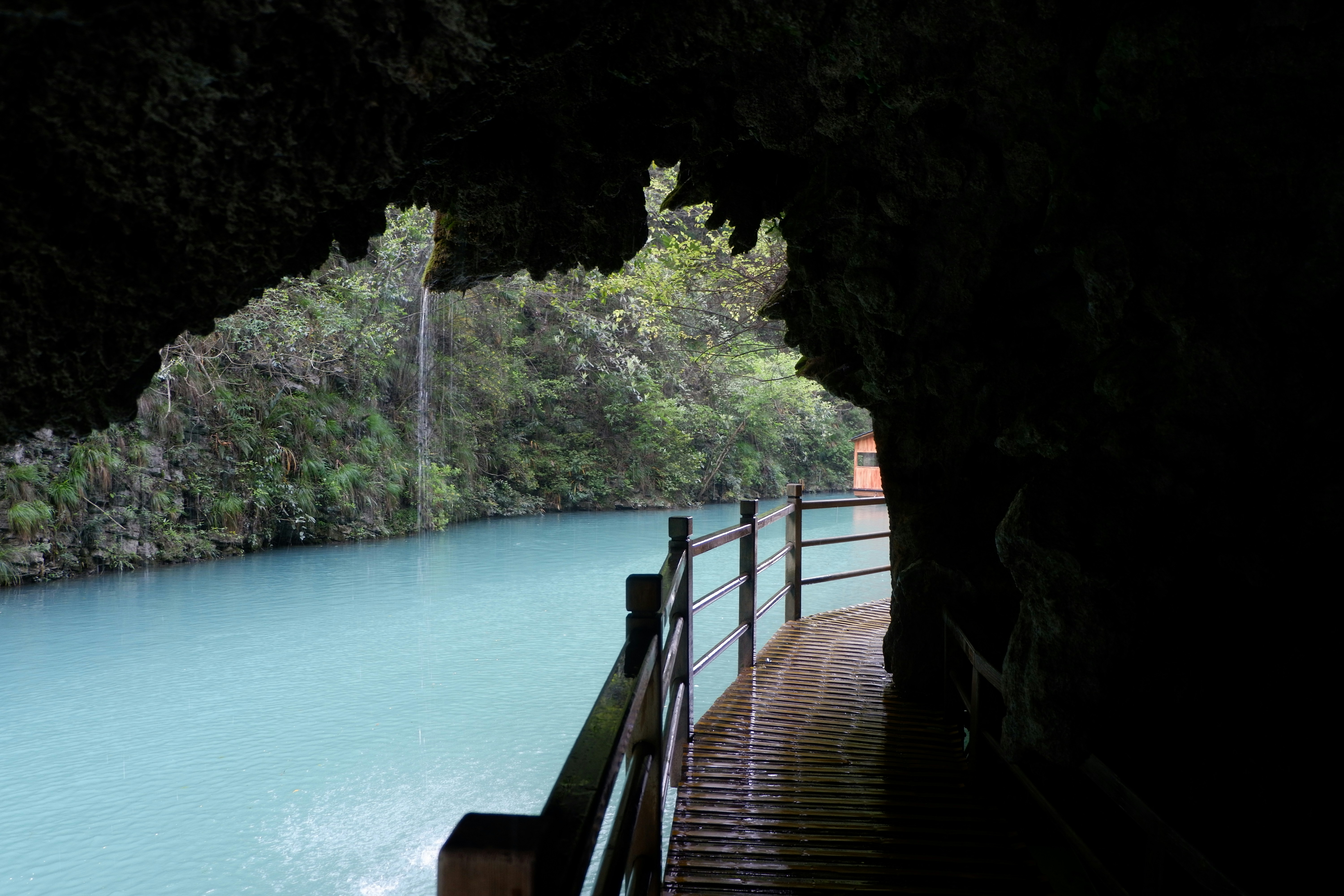 Wooden walkway through a cave opening to a tranquil turquoise river with lush greenery.