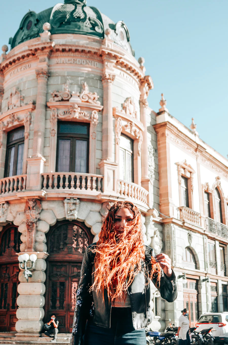Flame-haired woman standing before an ornate baroque theater façade with a domed roof under a clear pale-blue sky.