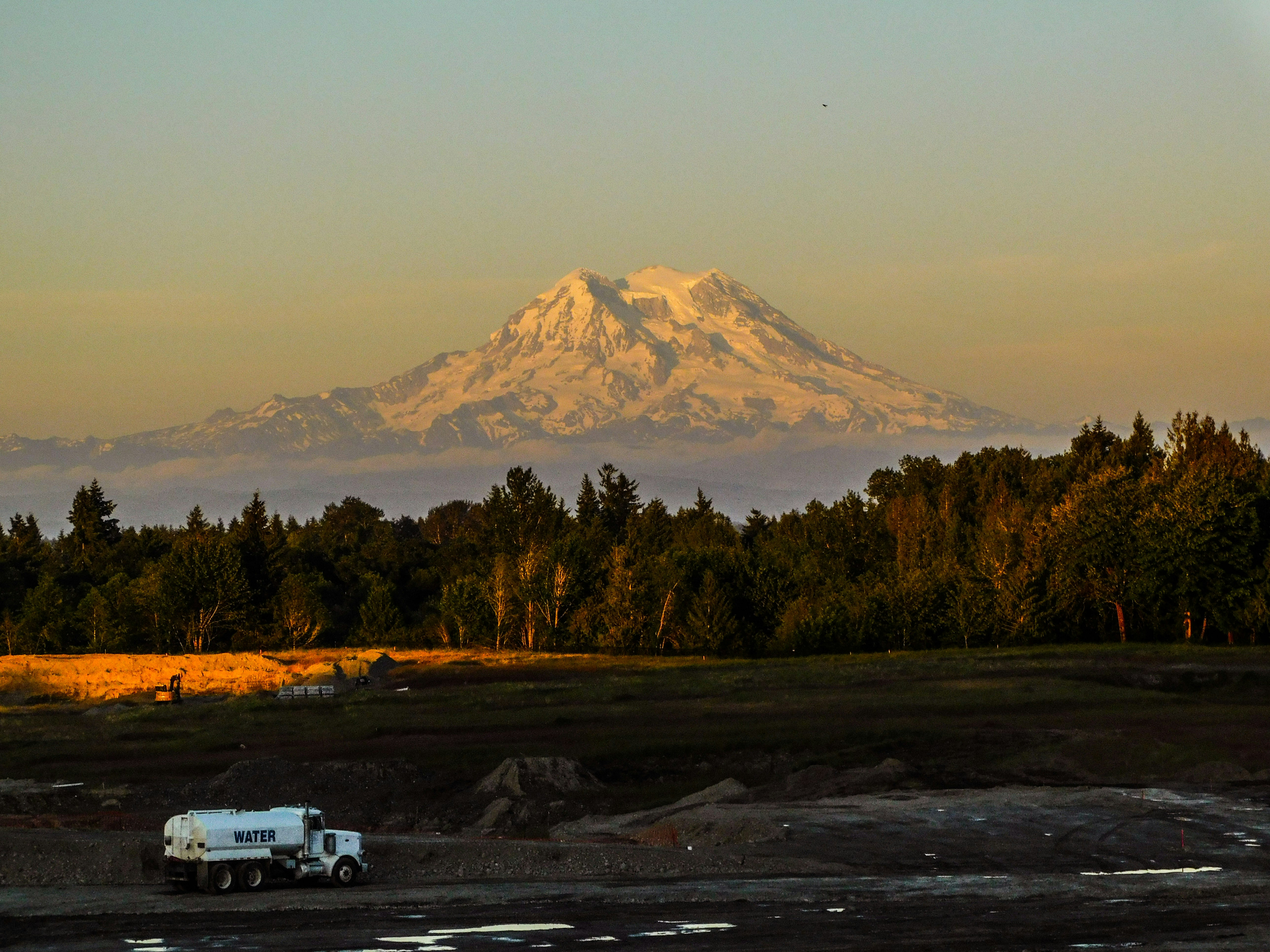 Golden-hour landscape featuring a snow-capped peak rising above a tree line. A dusty foreground with a small water truck anchors the scene.