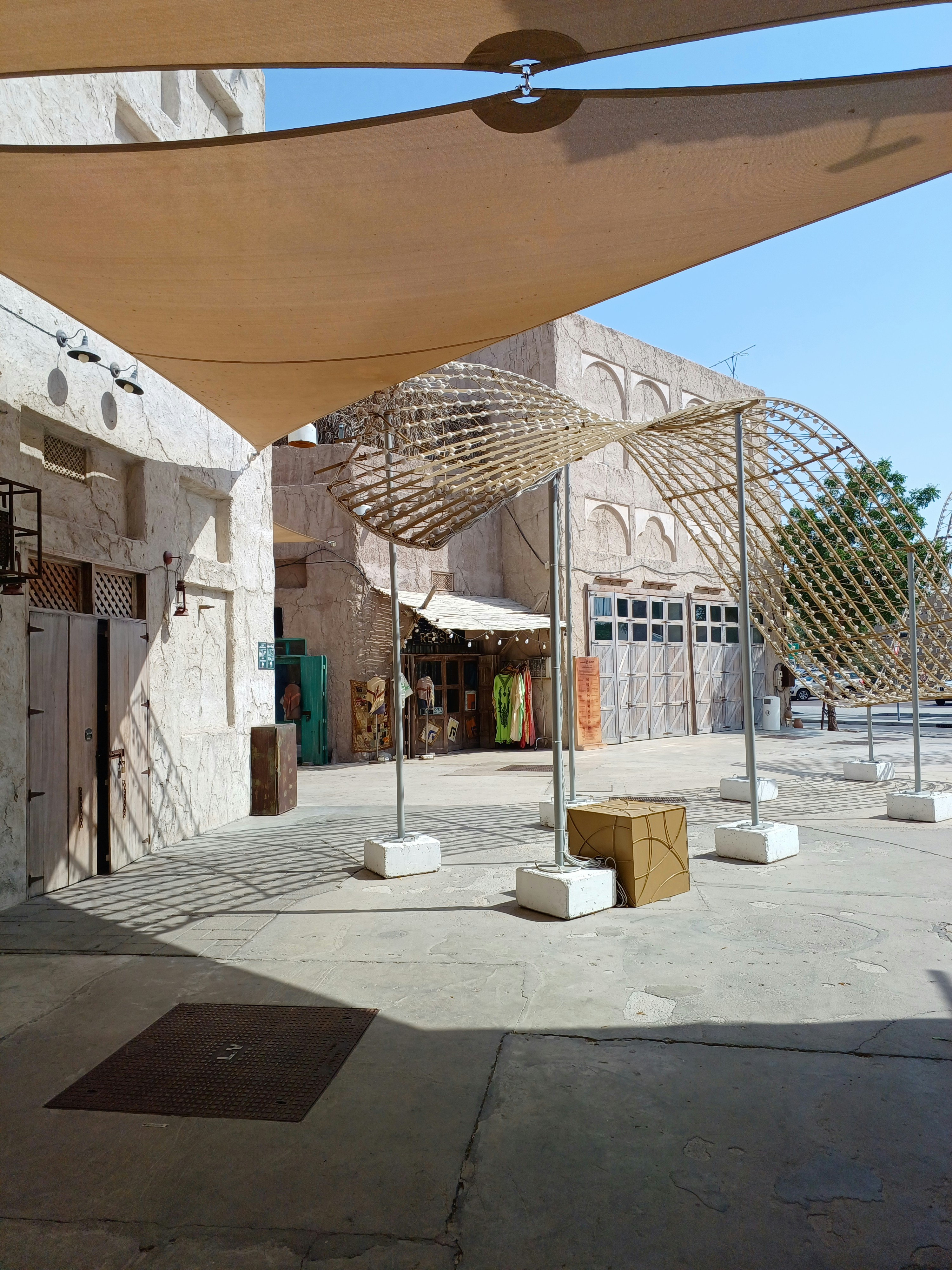 Sunlit courtyard framed by stone buildings, with large fabric shade sails overhead and a curved metal lattice canopy casting geometric shadows. Shoppers and stalls are visible in the background.