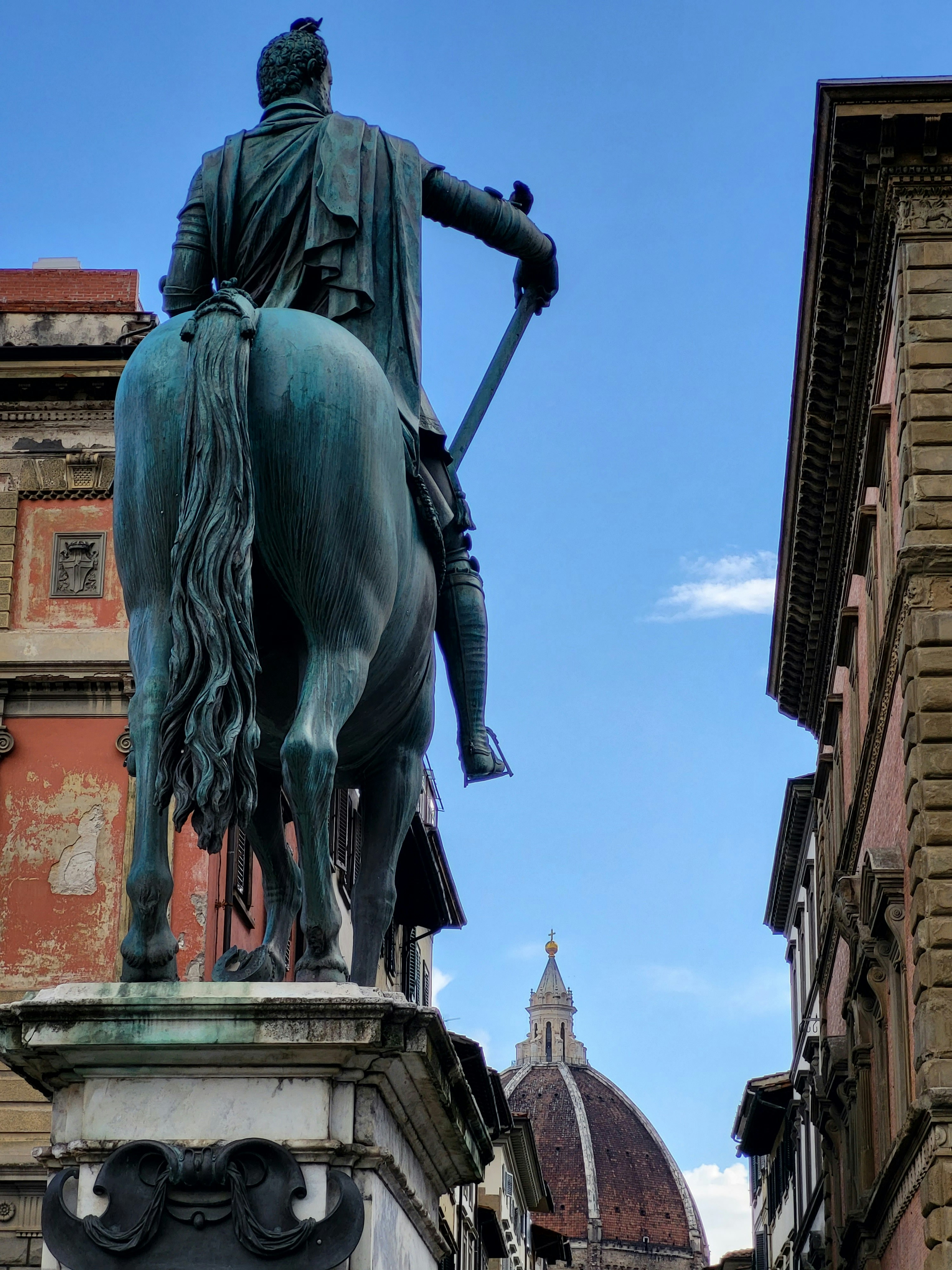 Bronze equestrian statue stands tall against a clear blue sky, with the iconic dome of Florence's cathedral visible in the background.