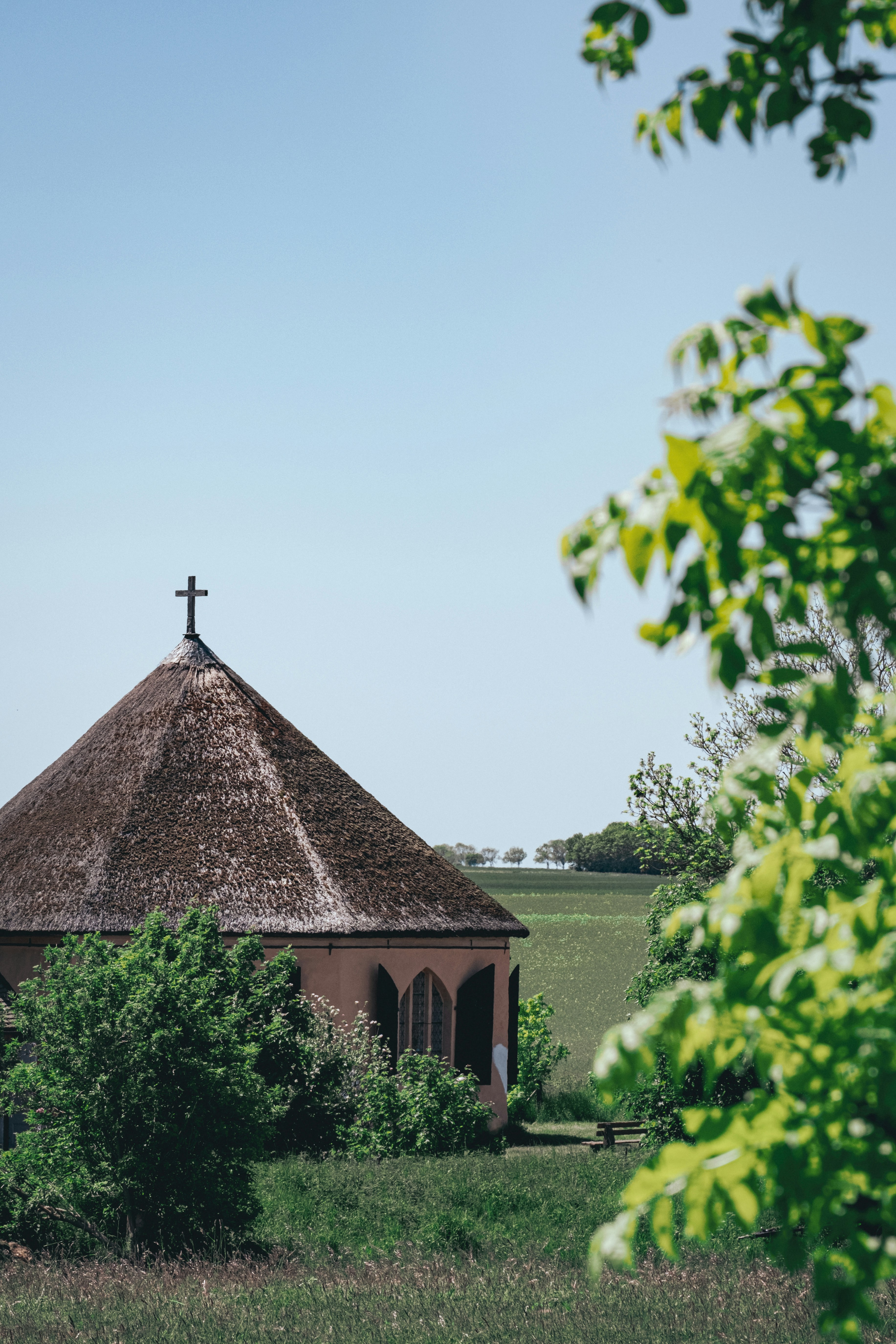 Thatched-roof chapel with a cross sits in a grassy field, framed by foreground greenery against a bright blue sky.