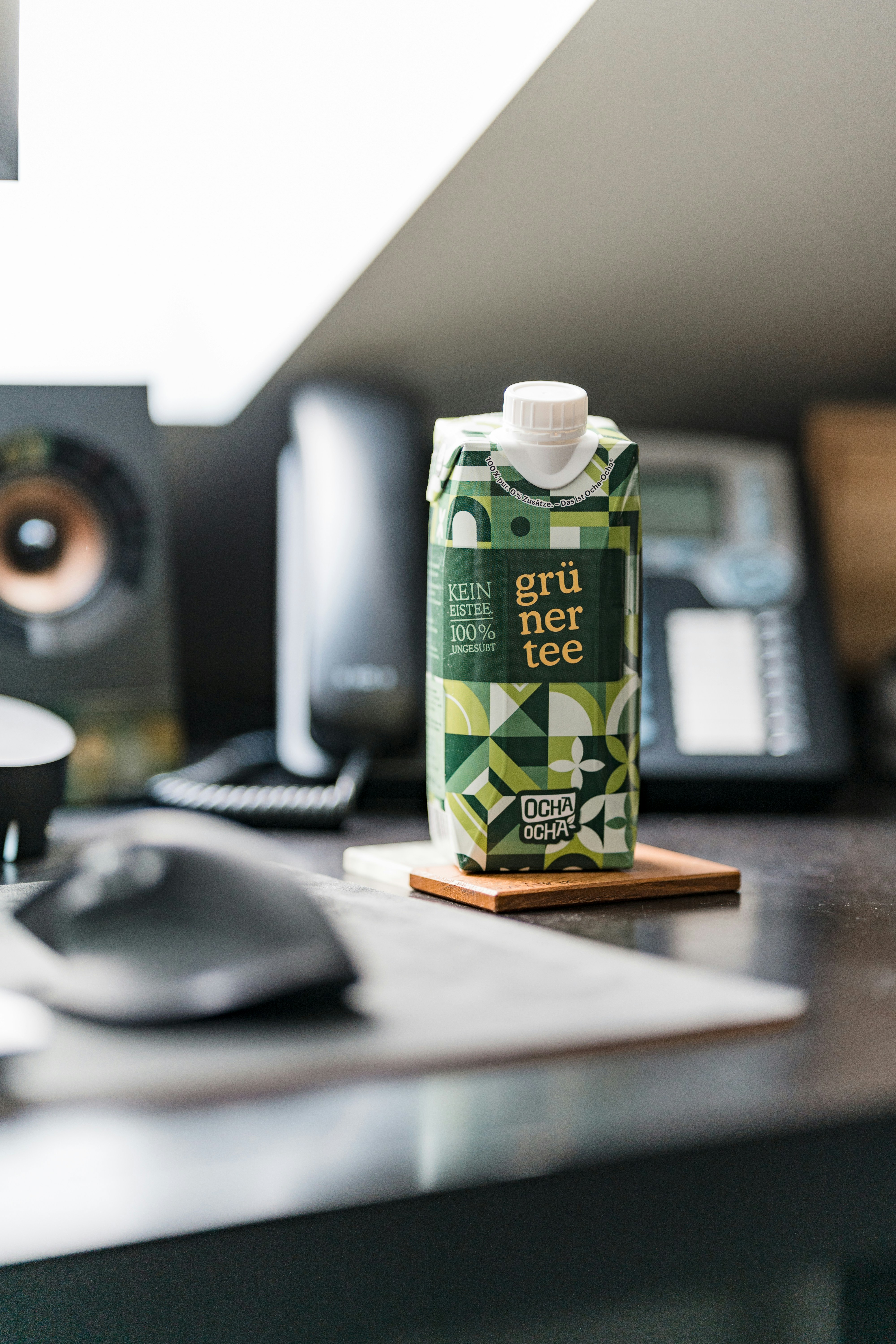 A green patterned juice carton sits on a wooden coaster atop a dark desk, with office equipment softly blurred in the background.