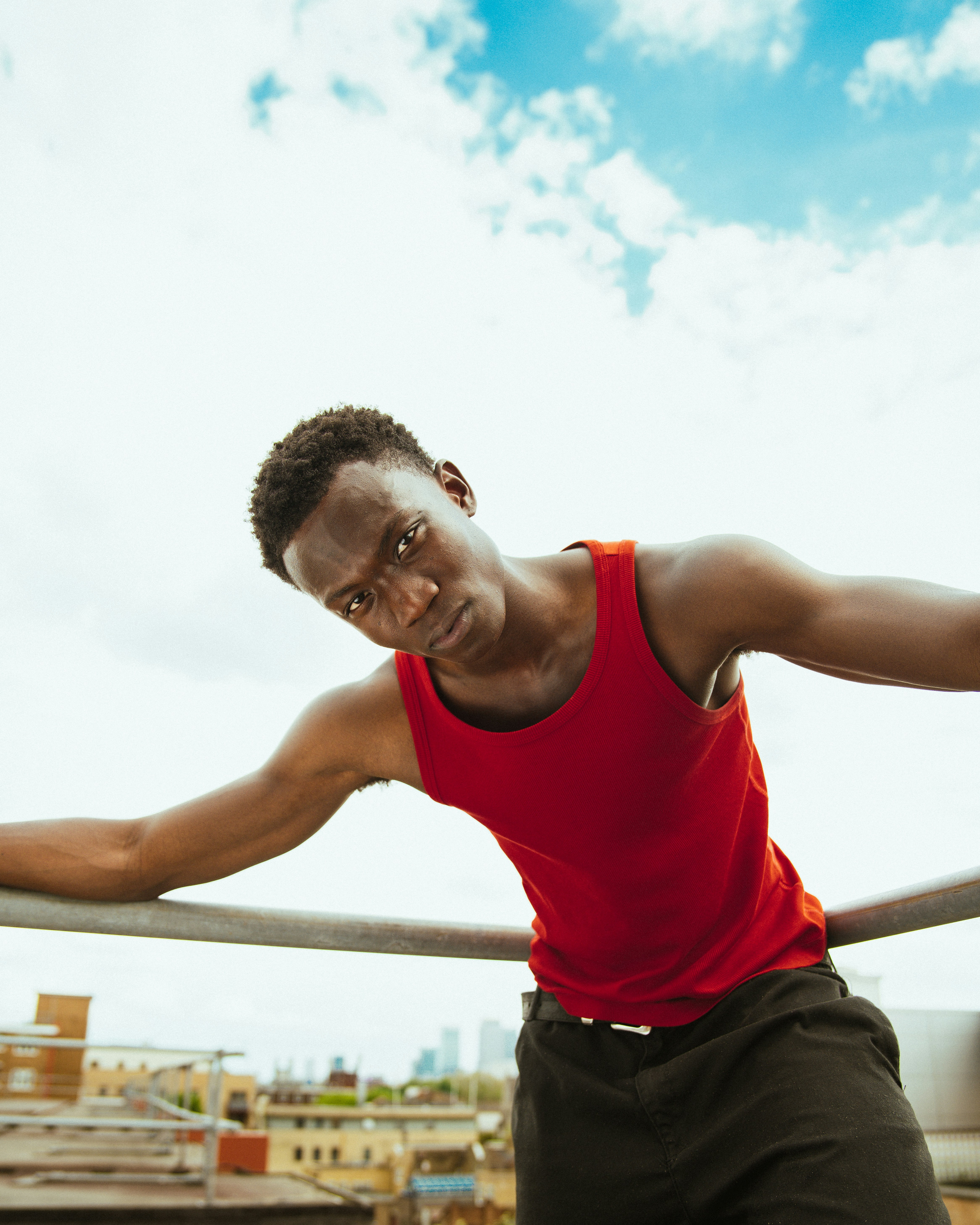 Young man in a red tank top leans against a railing on a rooftop, showcasing a confident pose against a backdrop of city buildings and blue sky.