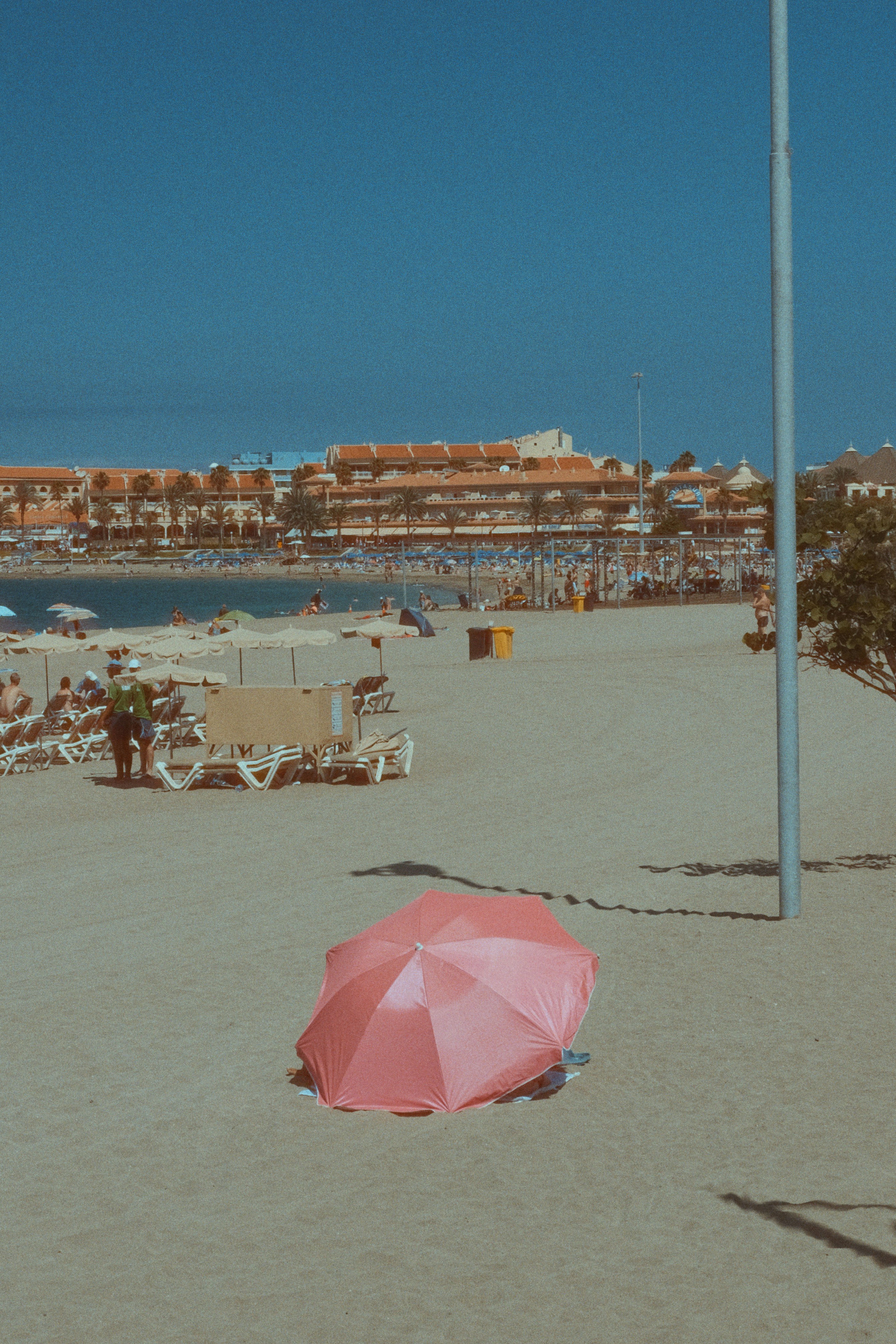 A pink umbrella sits alone on a sandy beach, surrounded by sun loungers and distant vacationers enjoying the vibrant seaside atmosphere.