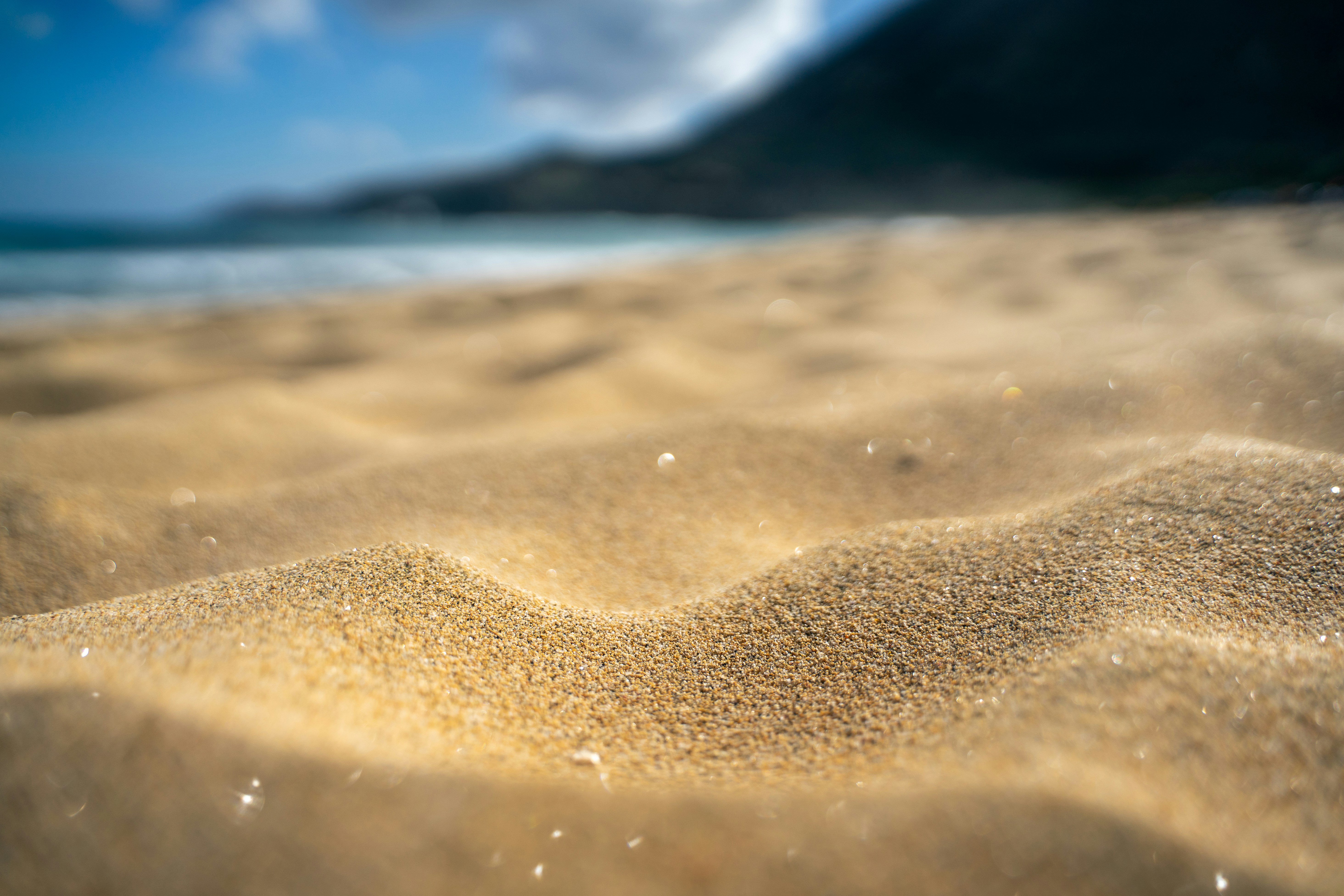 Close-up of rippled sand on a beach, capturing intricate patterns formed by the wind and waves. The blurred background hints at a serene coastal landscape.