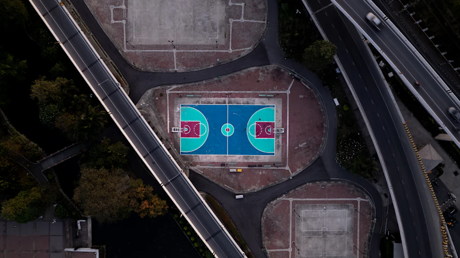 Top-down view of a bright blue and magenta basketball court surrounded by roads, overpasses, and dark trees. The geometric layout creates a striking urban pattern.