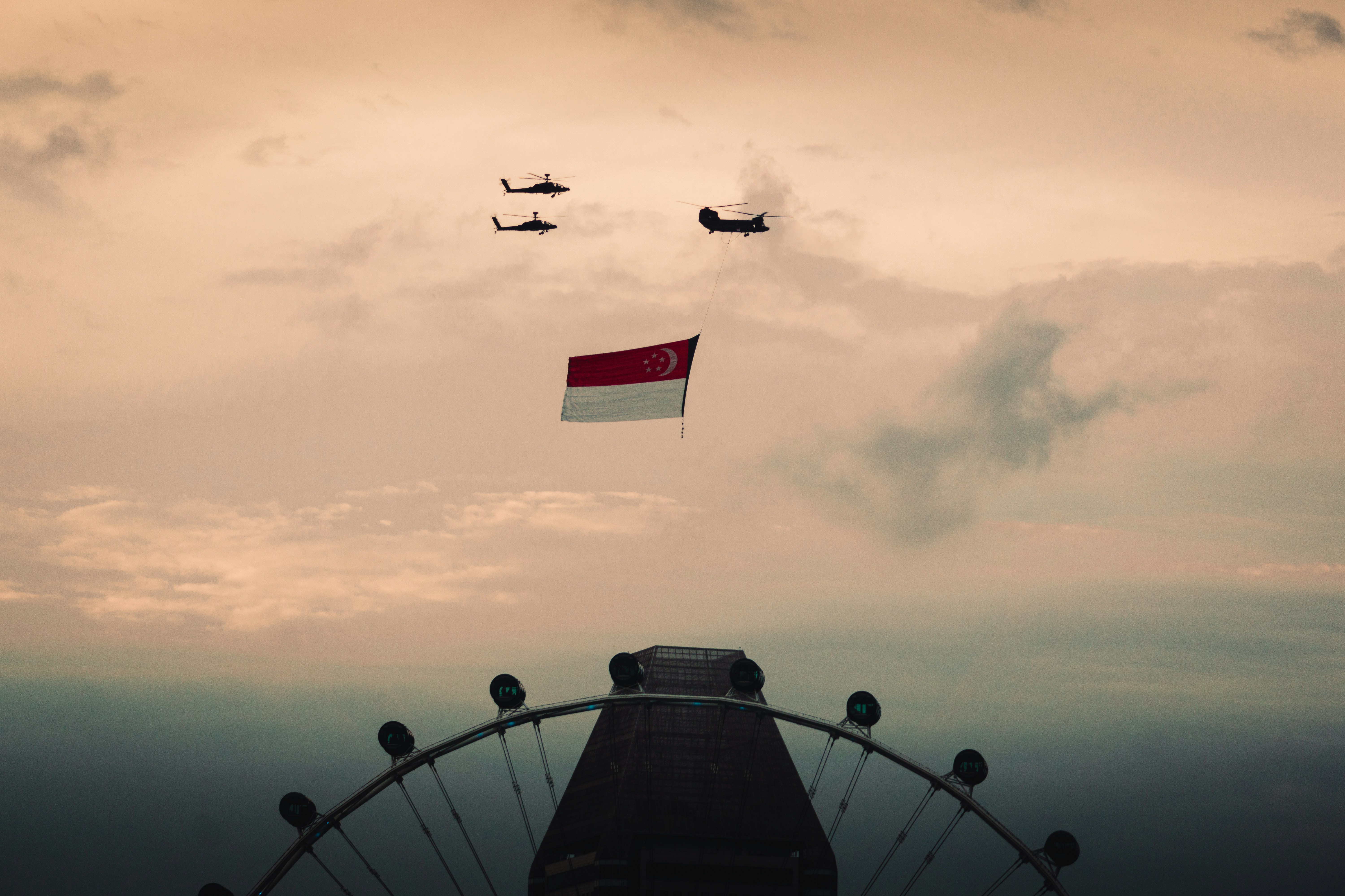 Singapore flag billowing in the wind as helicopters fly overhead, framed by a giant Ferris wheel against a moody sky.
