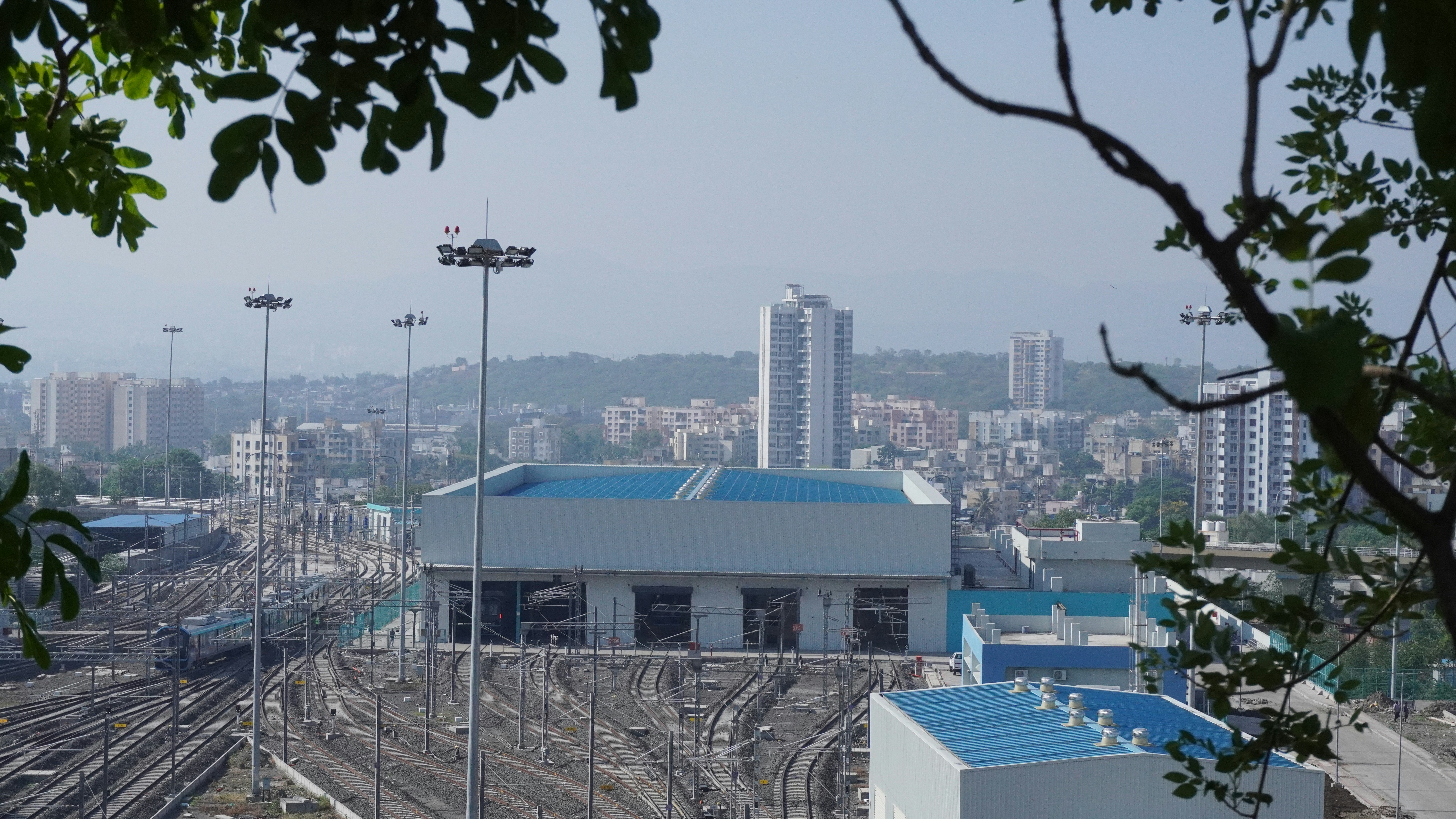 A sprawling railway yard with intricate tracks and modern buildings, framed by lush foliage in the foreground.