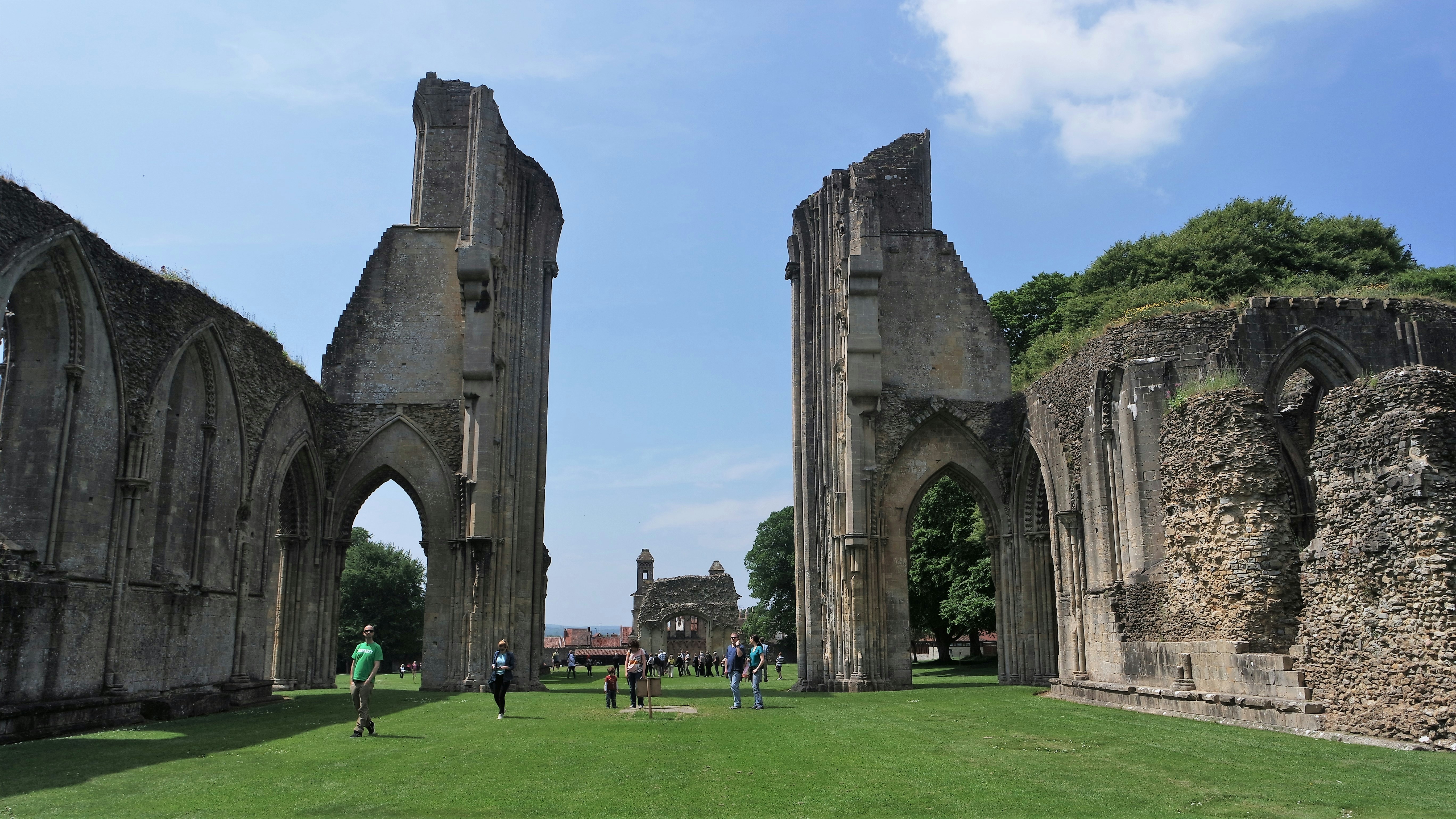 Ancient ruin arches rise beside a manicured lawn, as visitors explore the sunlit site under a clear blue sky.