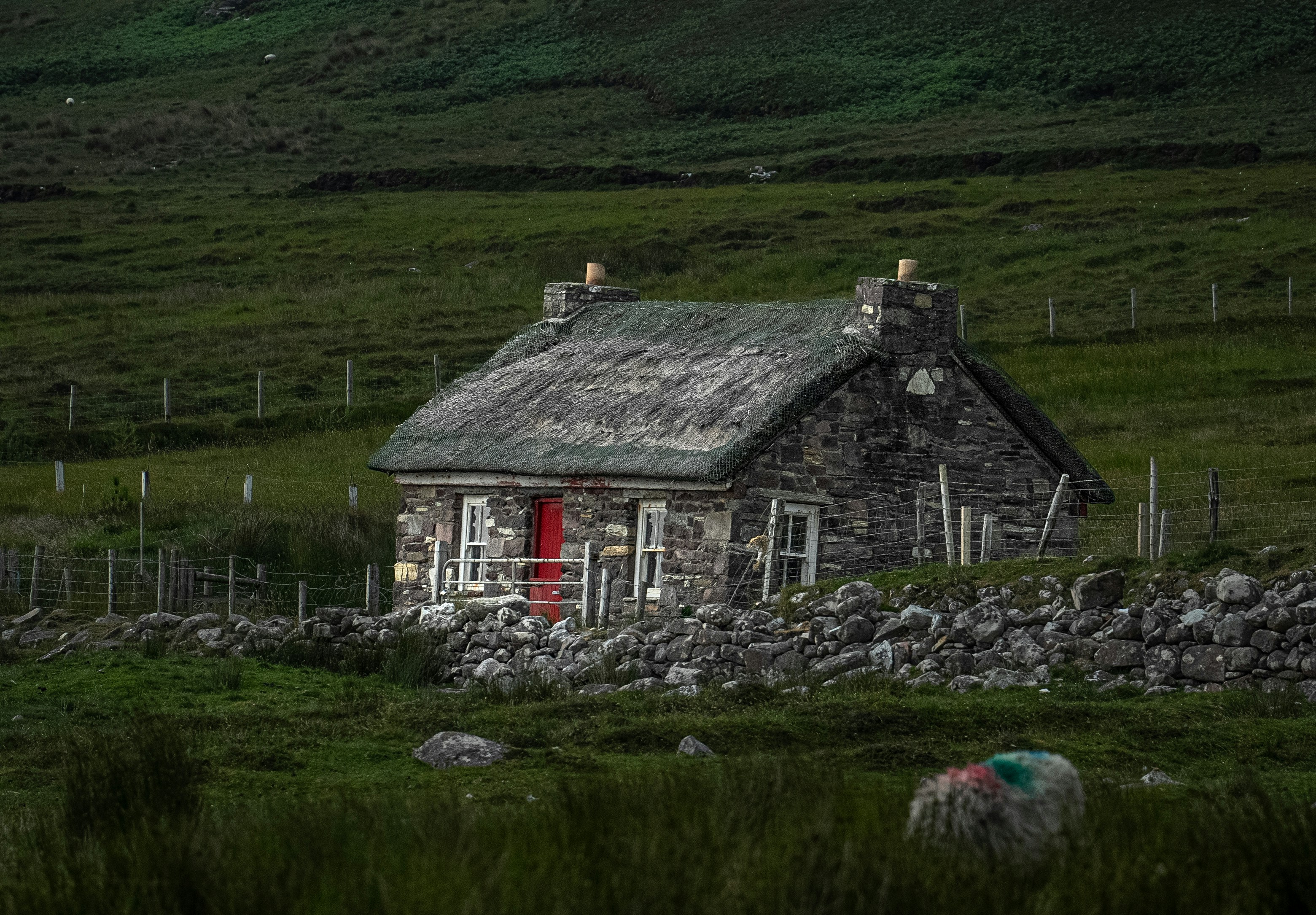 A quaint stone cottage with a vibrant red door stands alone in a lush green landscape, surrounded by a rustic stone wall.