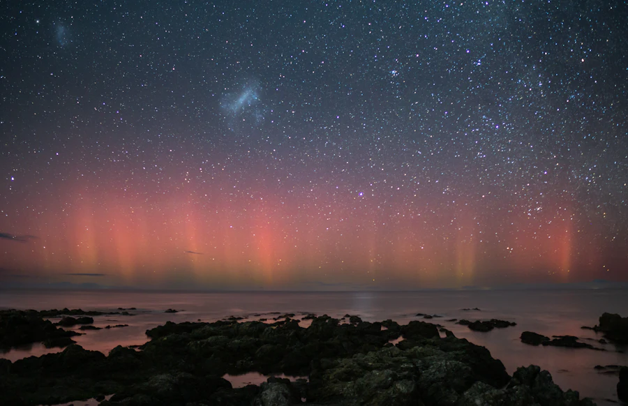 Pink-orange aurora curtains shimmer beneath a star-filled night sky above dark rocky shoreline and calm water.