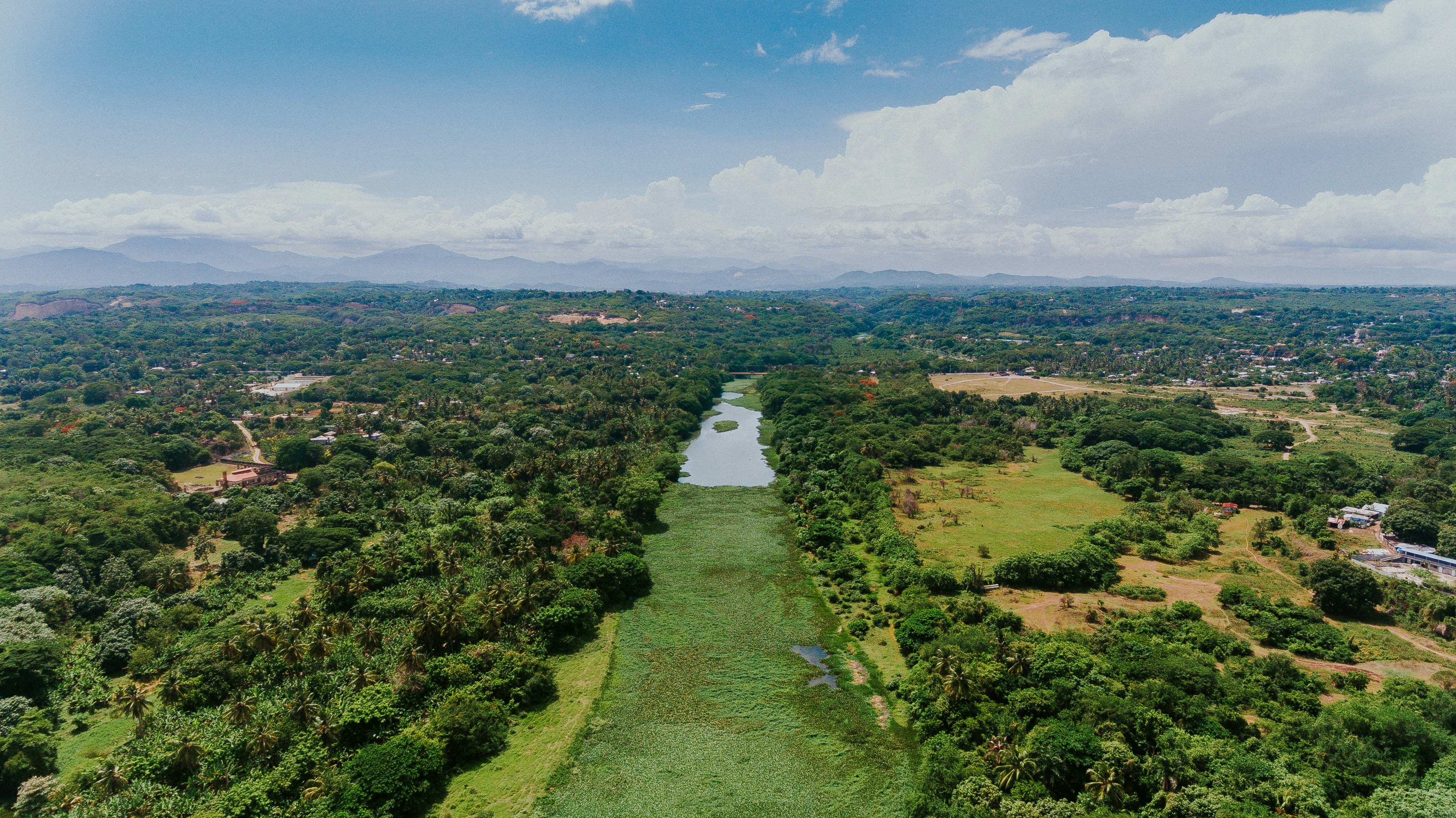 Expansive aerial view of a lush green landscape with a river running through fields under a partly cloudy sky.