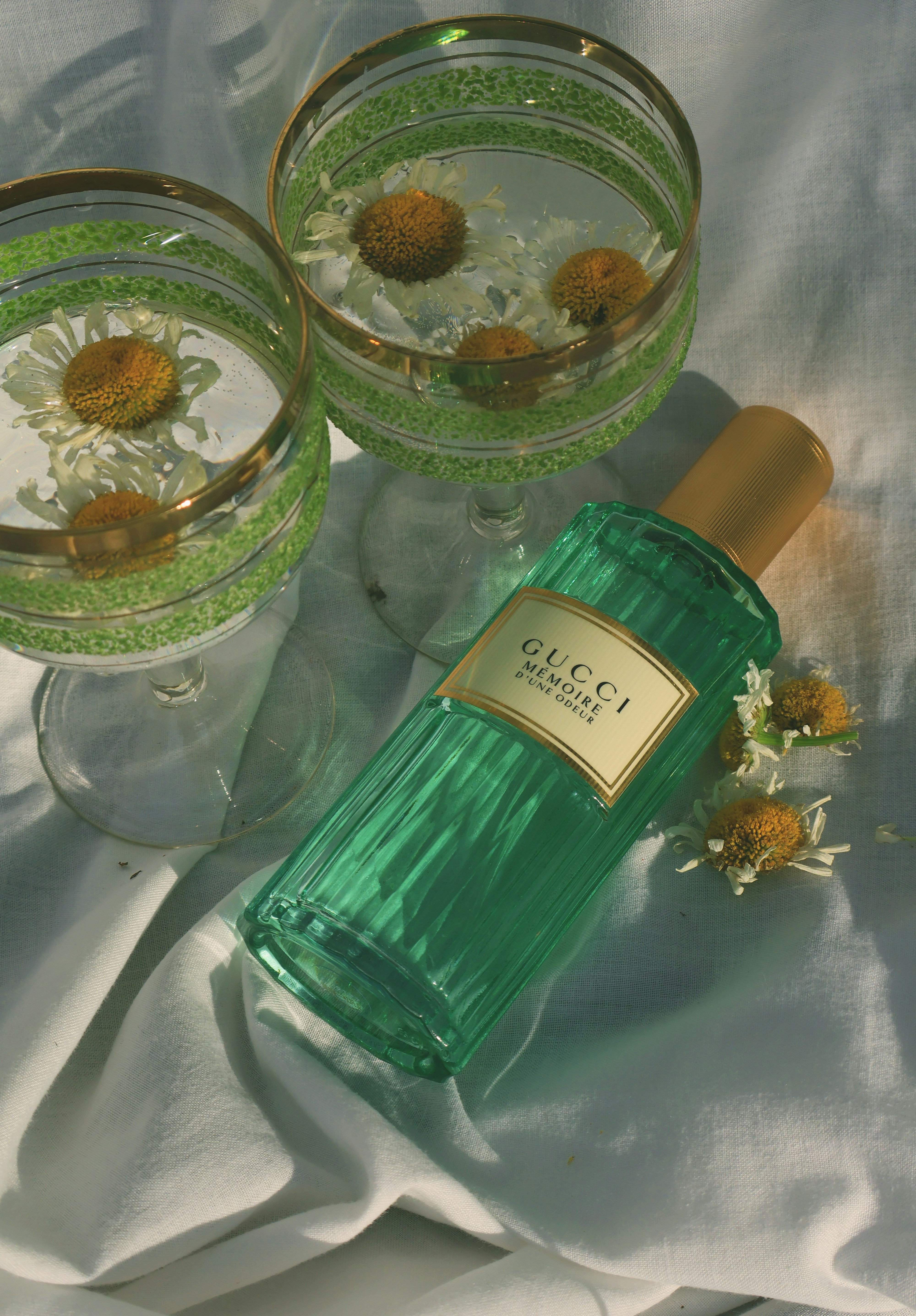 Still-life photograph of a Gucci perfume bottle in emerald glass resting beside two daisy-filled champagne glasses on white fabric.