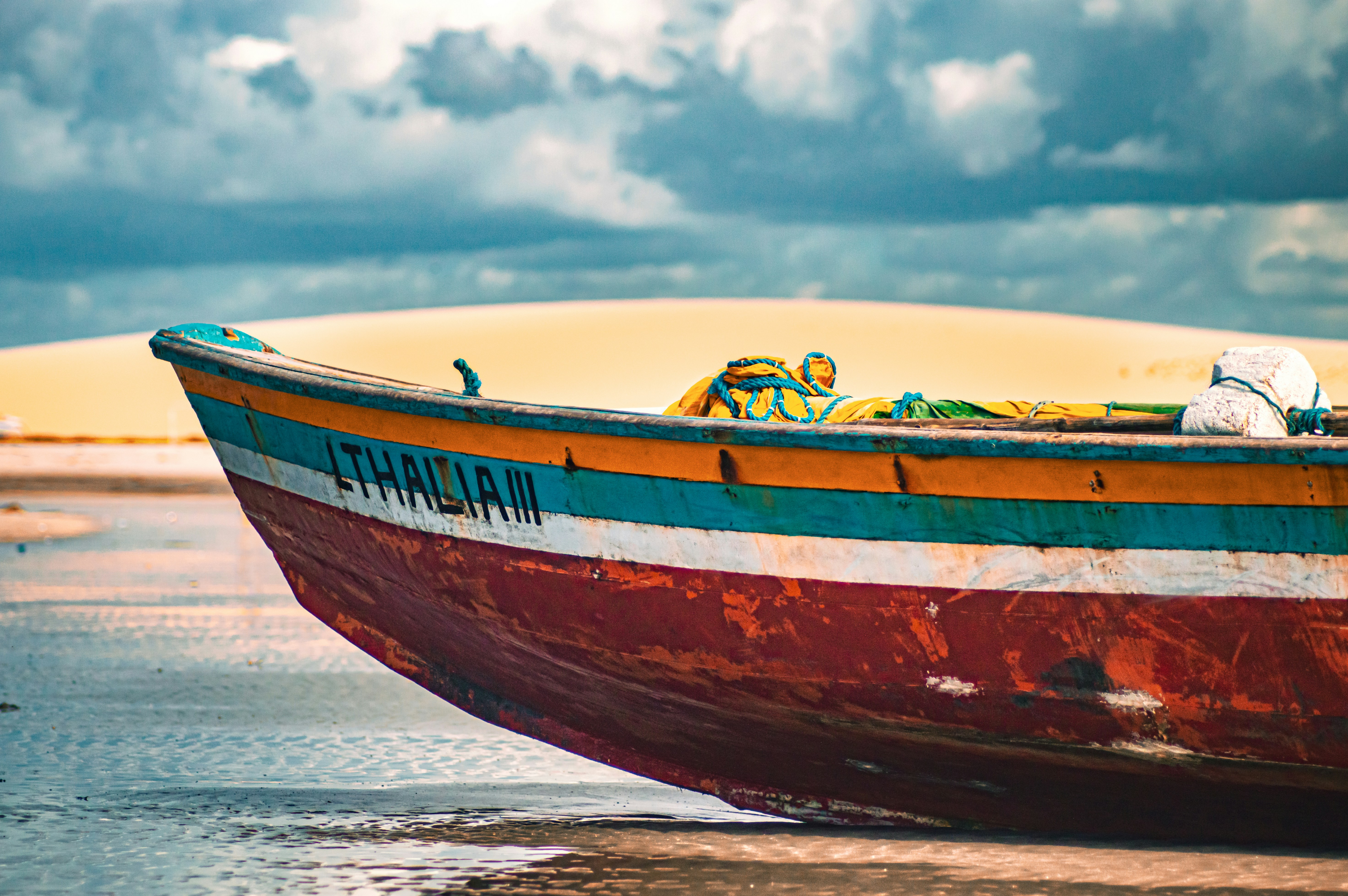Colorful fishing boat resting on the shore with a backdrop of rolling sand dunes and dramatic clouds.