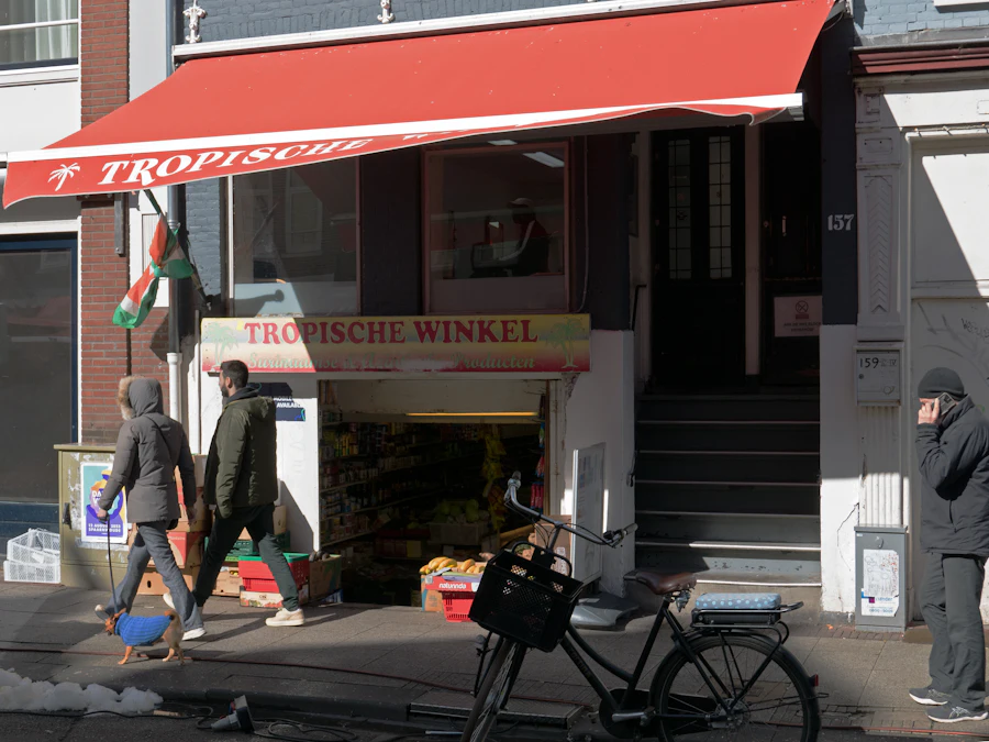 Street-side shopfront with a red awning reading “TROPISO…” and a lower sign for “TROPISCHE WINKEL.” Pedestrians pass by a bicycle parked in front of the entrance.