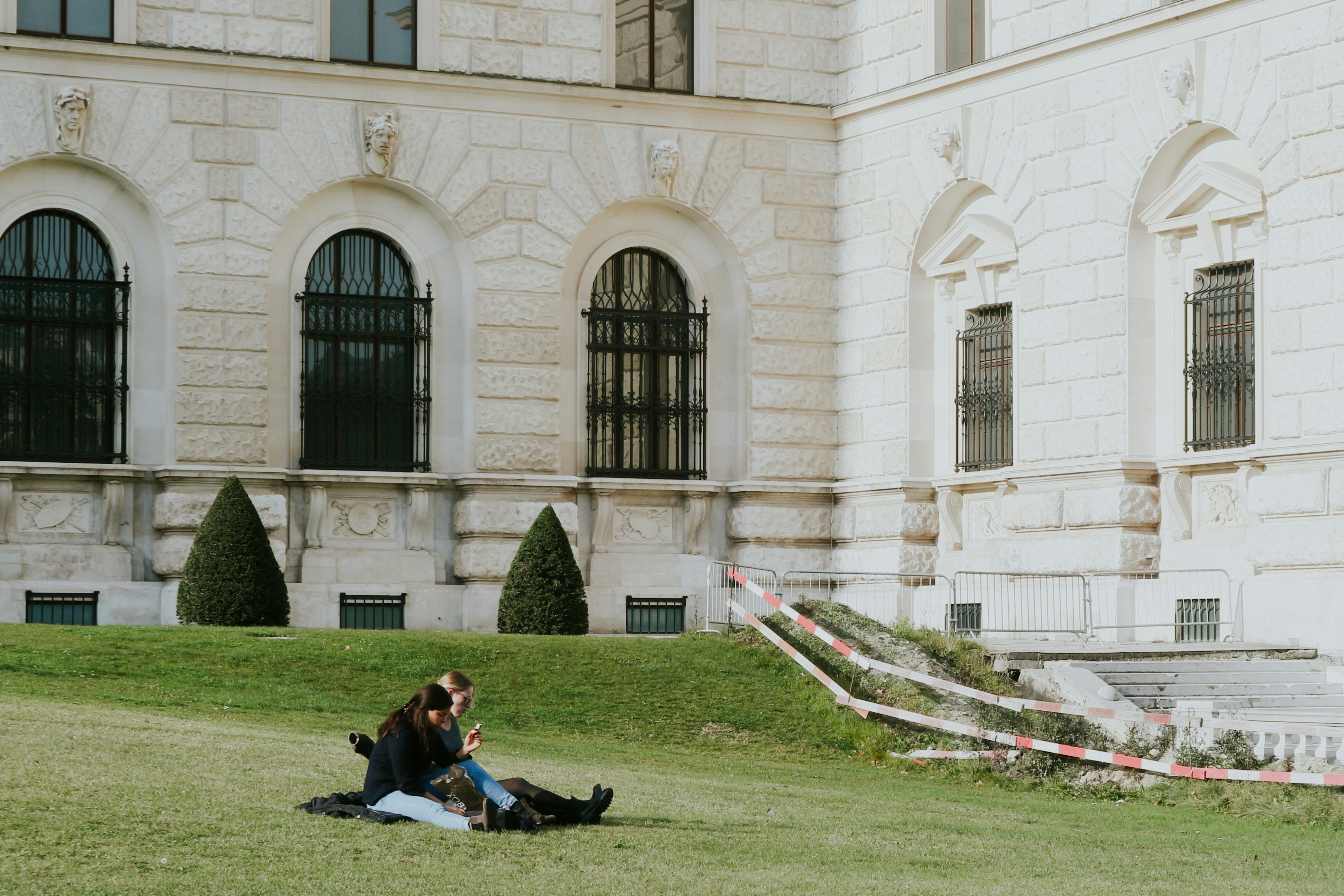 Two individuals seated on a grassy area, engaged with their devices, against a backdrop of an ornate building with large windows and manicured shrubs.