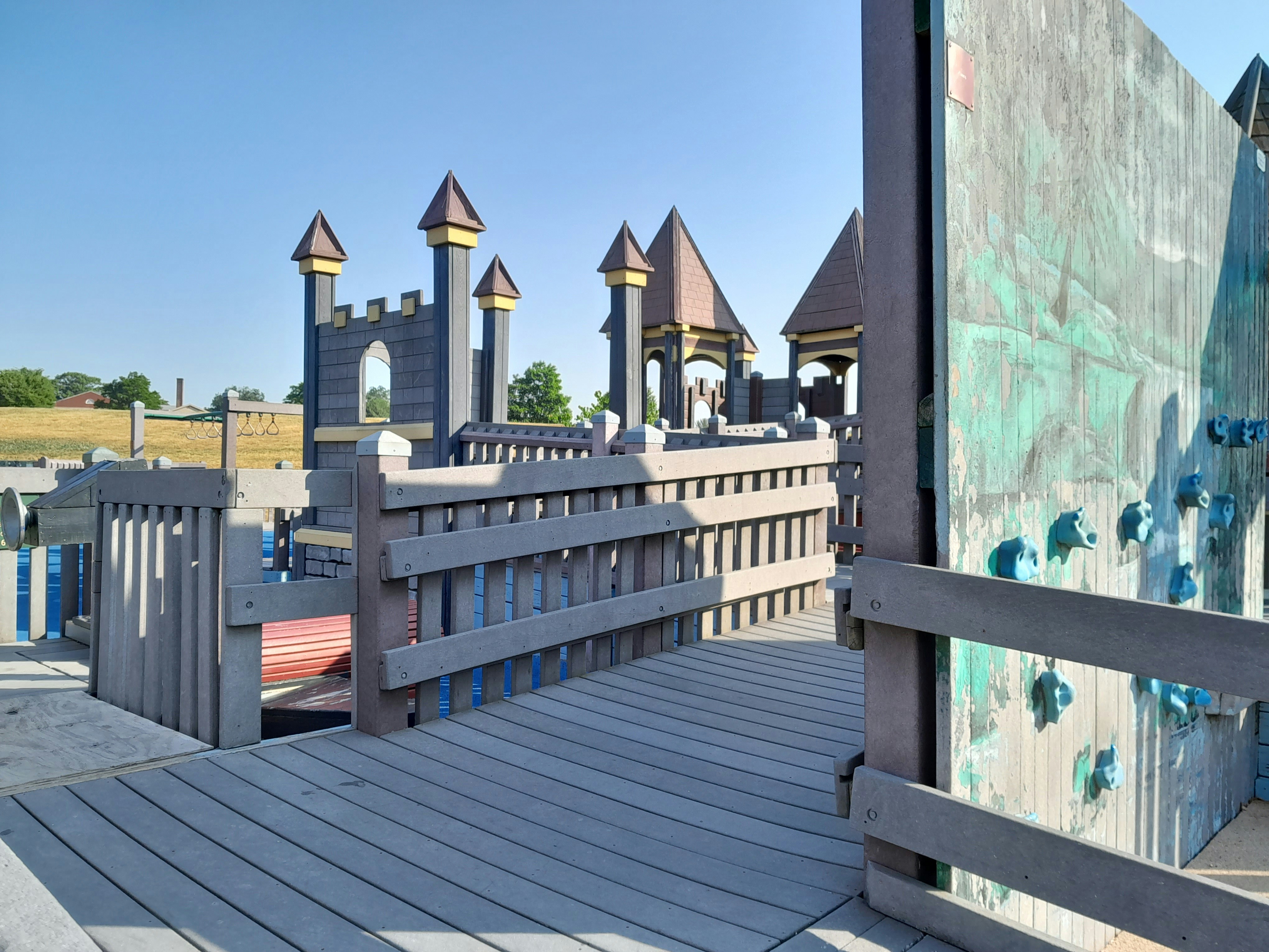 Wooden castle-style playground with towers and railings sits along a sunlit boardwalk under a clear blue sky.