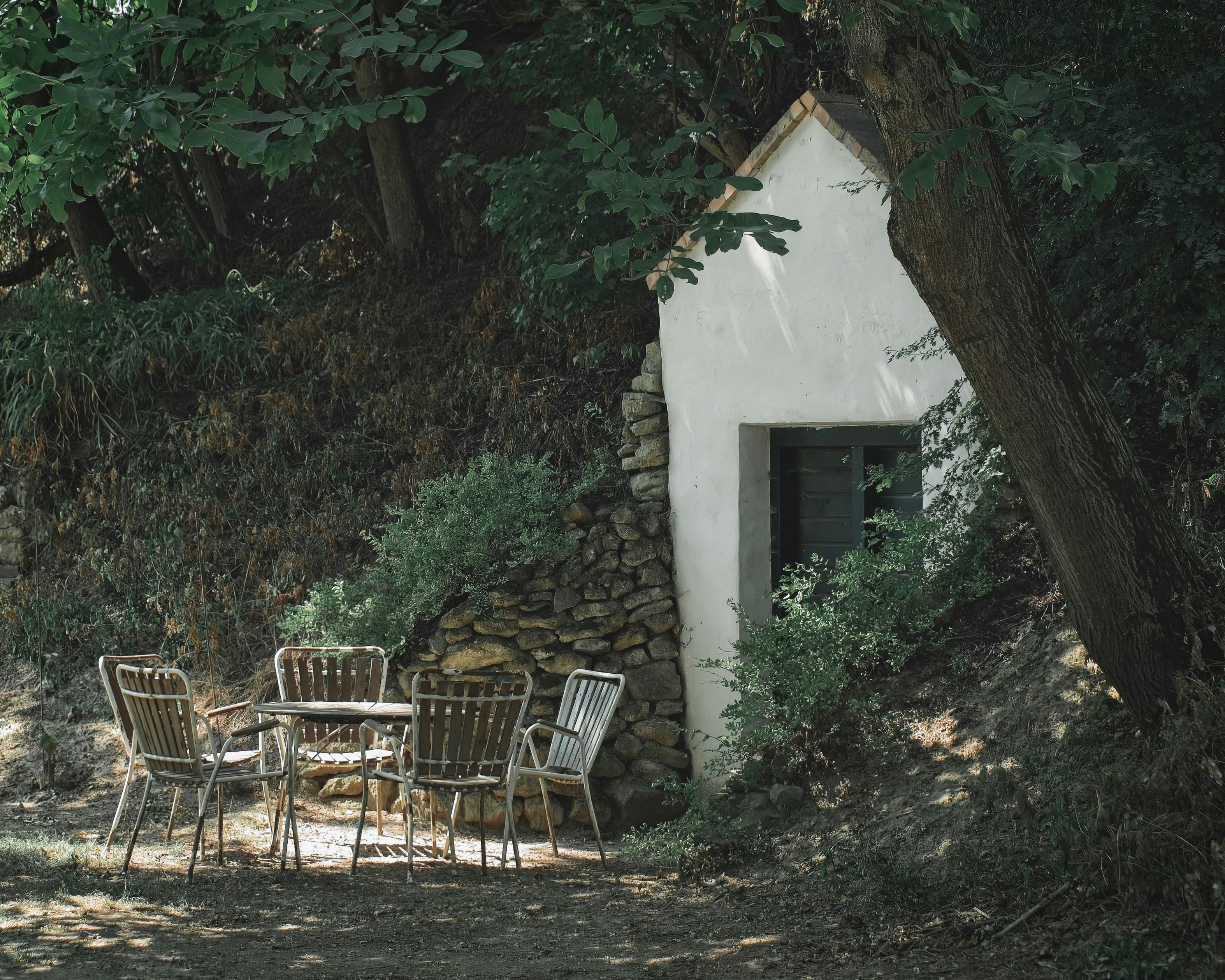 Outdoor seating area with metal chairs arranged around a round table beside a small stone-walled shed, tucked under overhanging trees.