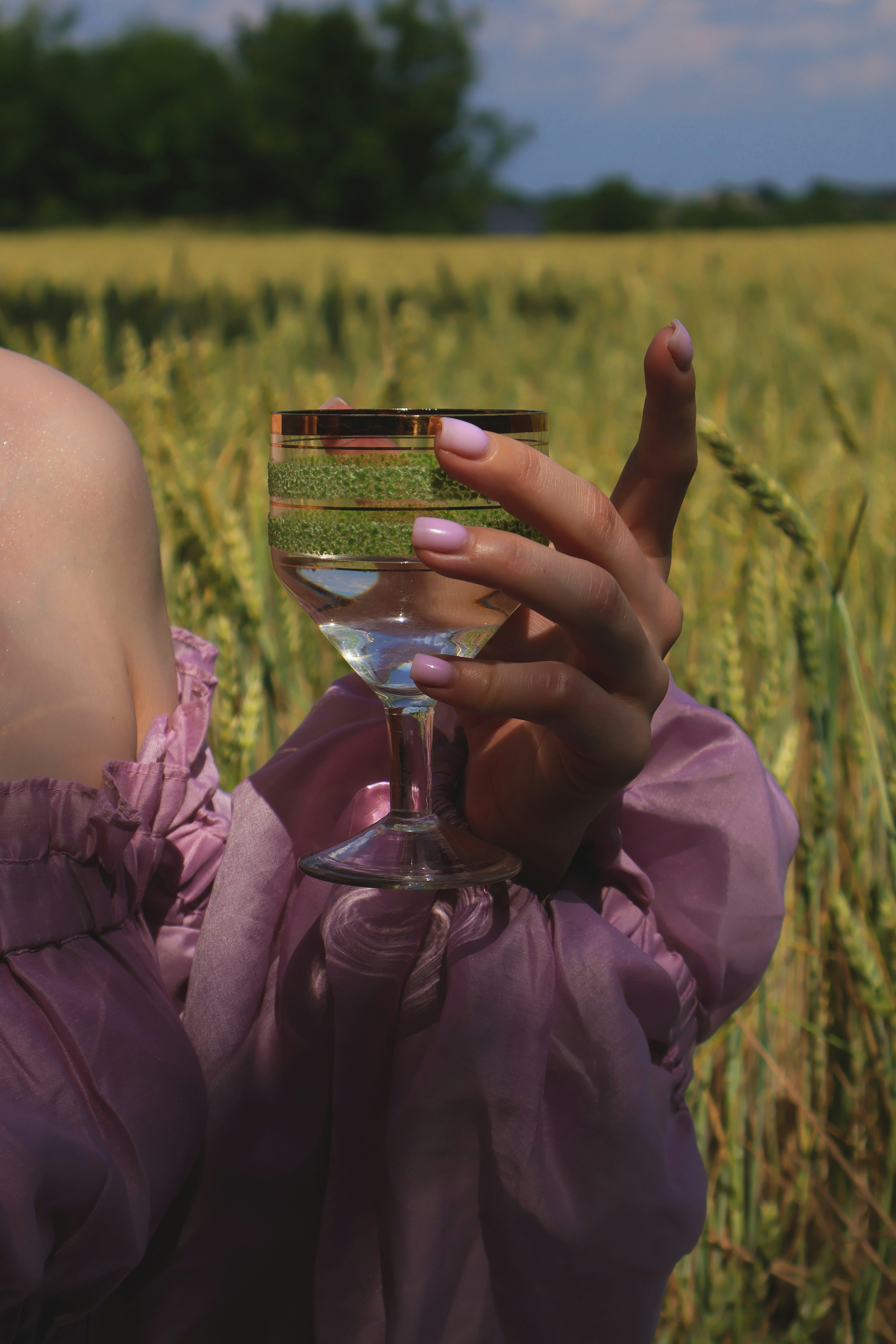 A hand in a lavender off-shoulder blouse holds a crystal glass of water in a sunlit field of tall, golden crops.