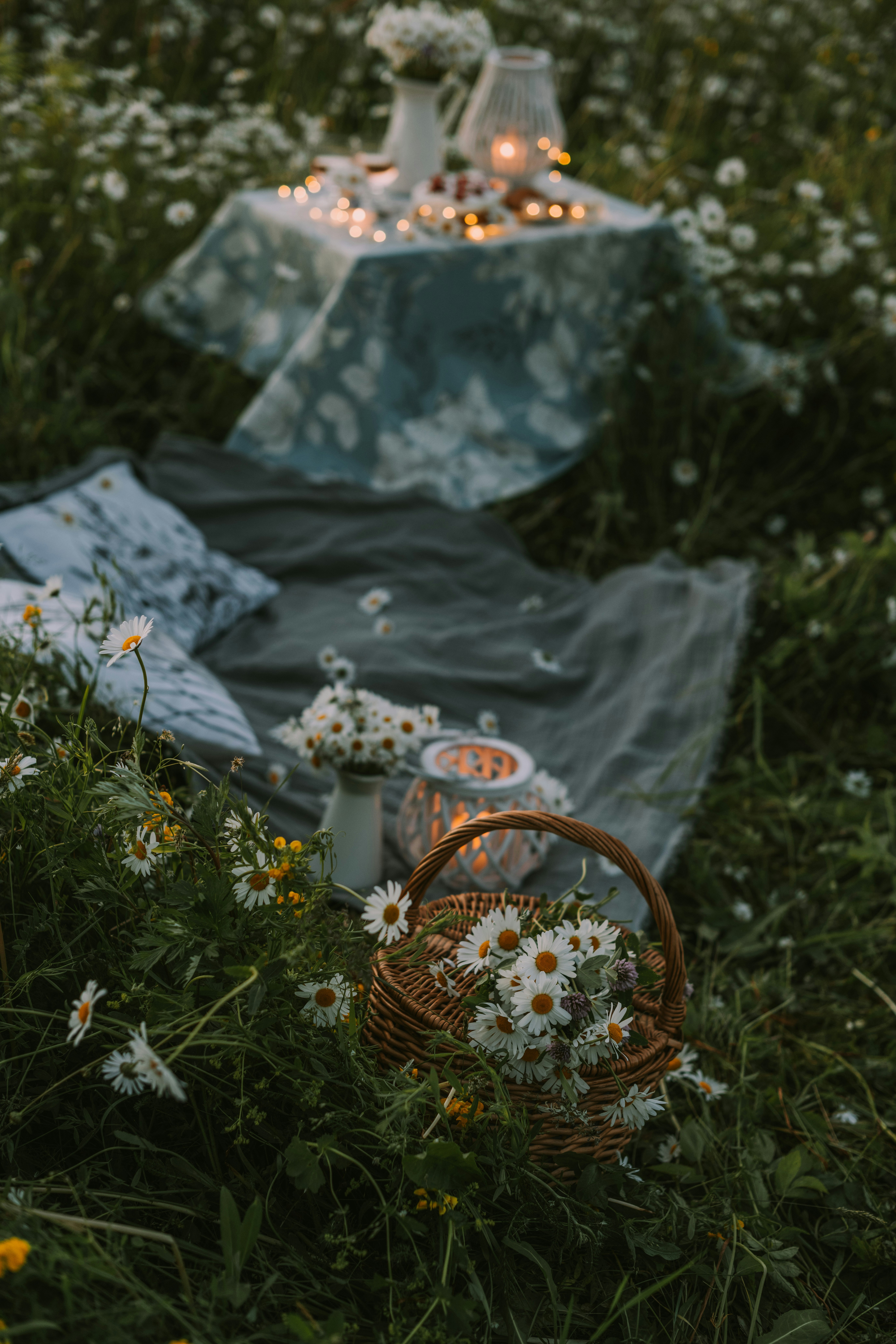 Softly lit meadow picnic photo featuring a wicker basket of daisies, a candlelit table on a blue cloth, and wildflowers.