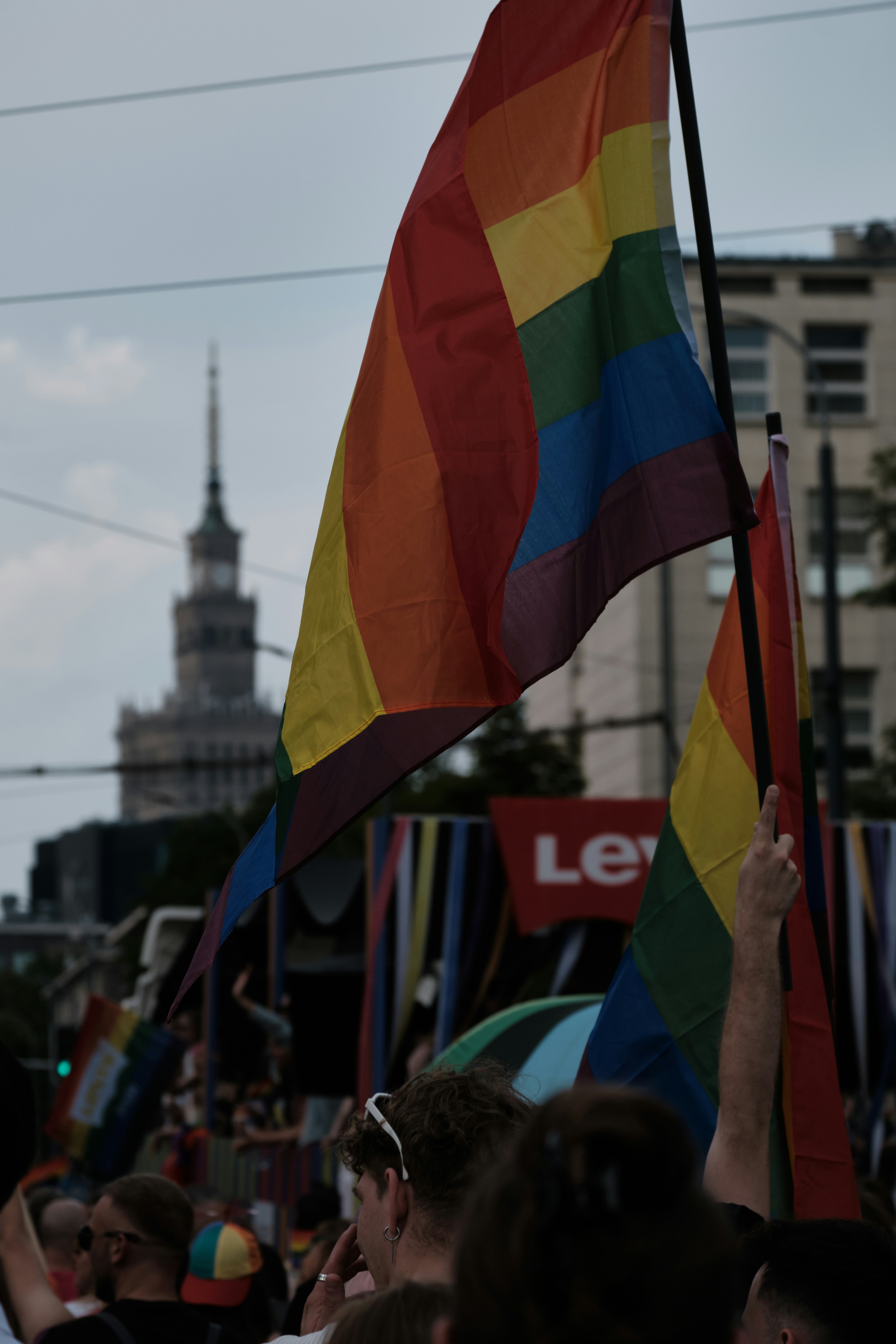 Vibrant Pride flags ripple over a densely packed crowd along a city street, with a historic clocktower rising in the background.