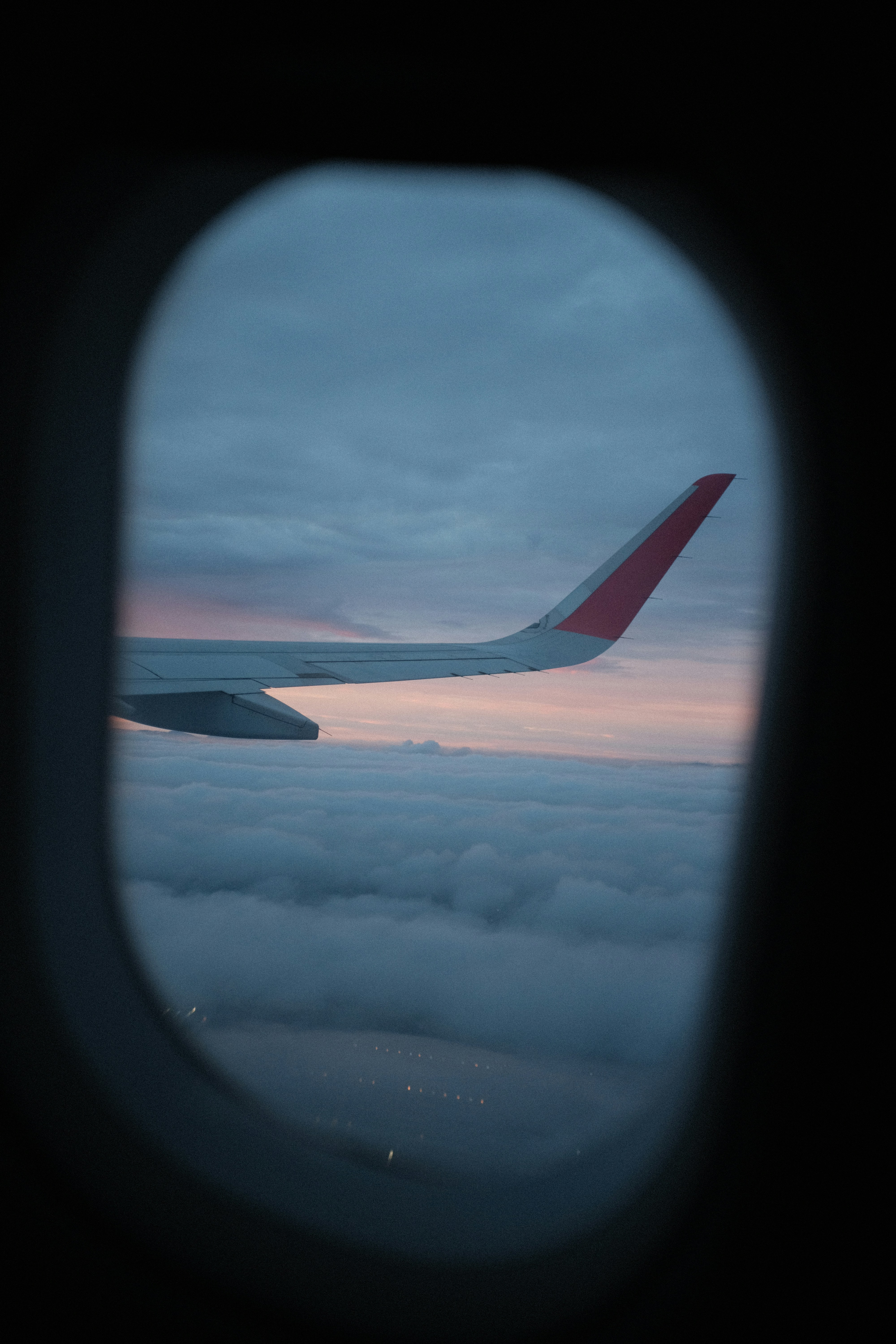 Airplane wing extending into a sky painted with soft hues, framed by an aircraft window. The scene captures the tranquility of flight amidst a sea of clouds.