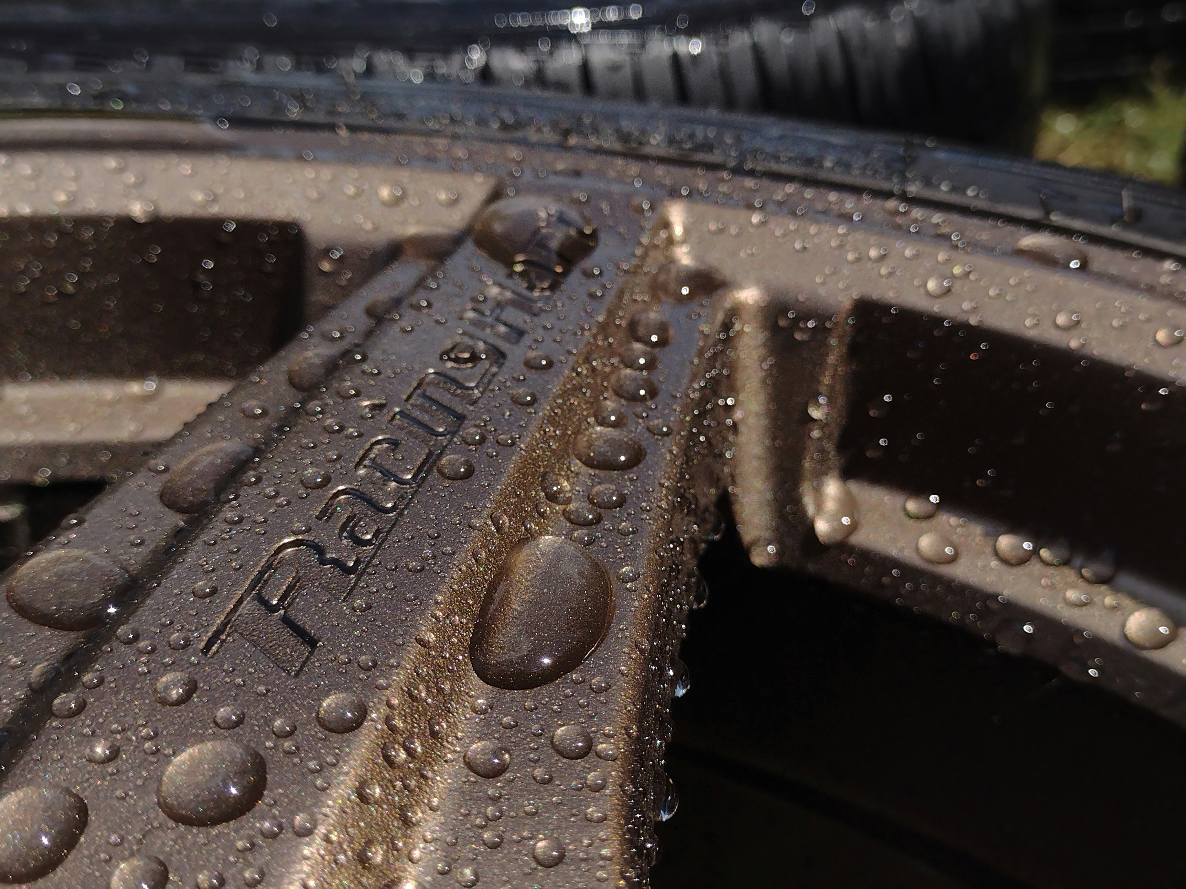 Macro photograph of a rain-speckled car wheel rim, highlighting texture and liquid beads along the metallic spokes.
