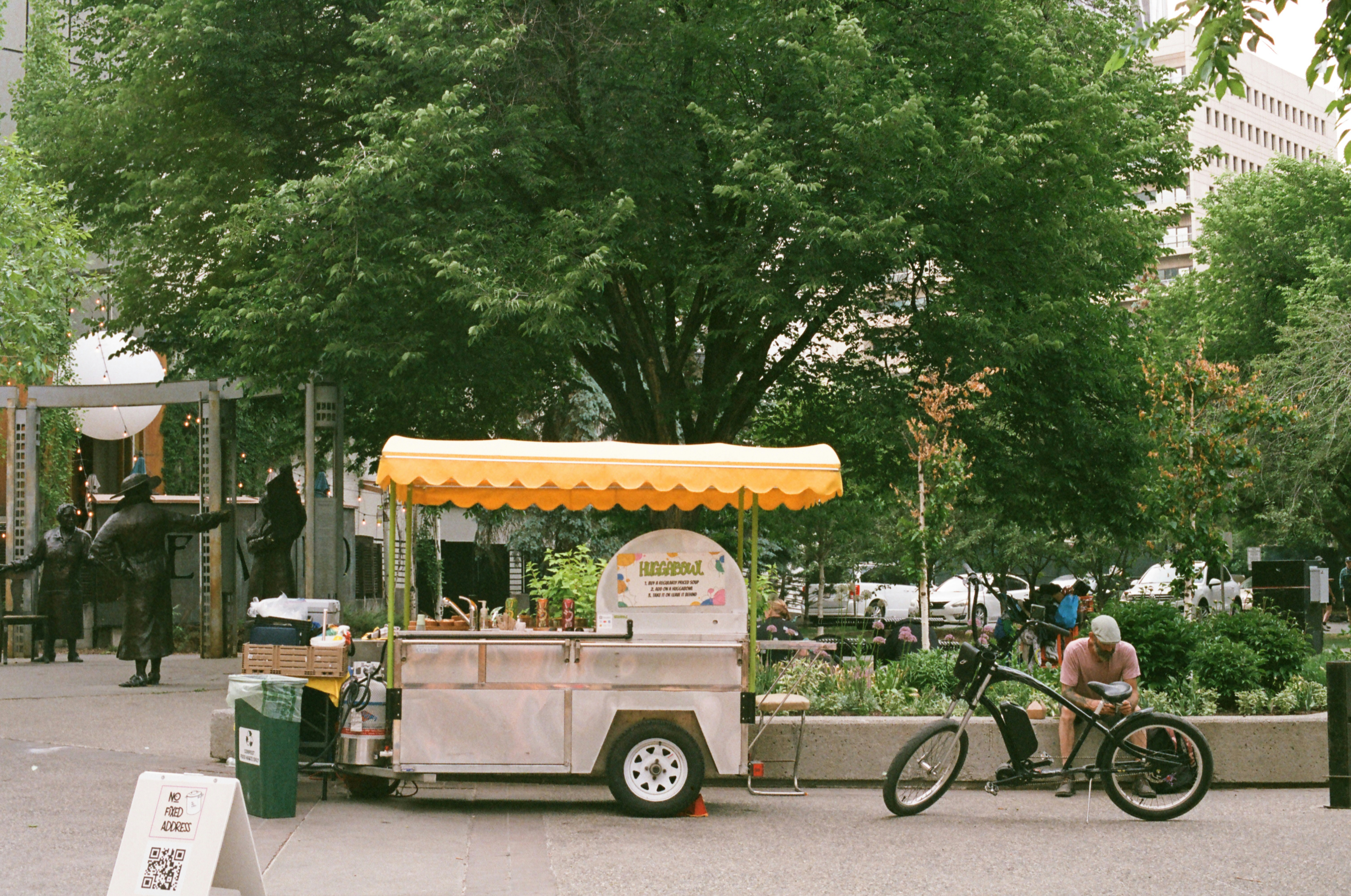 City park photograph of a yellow-canopy food cart on a plaza, with a bicycle nearby and green trees overhead.