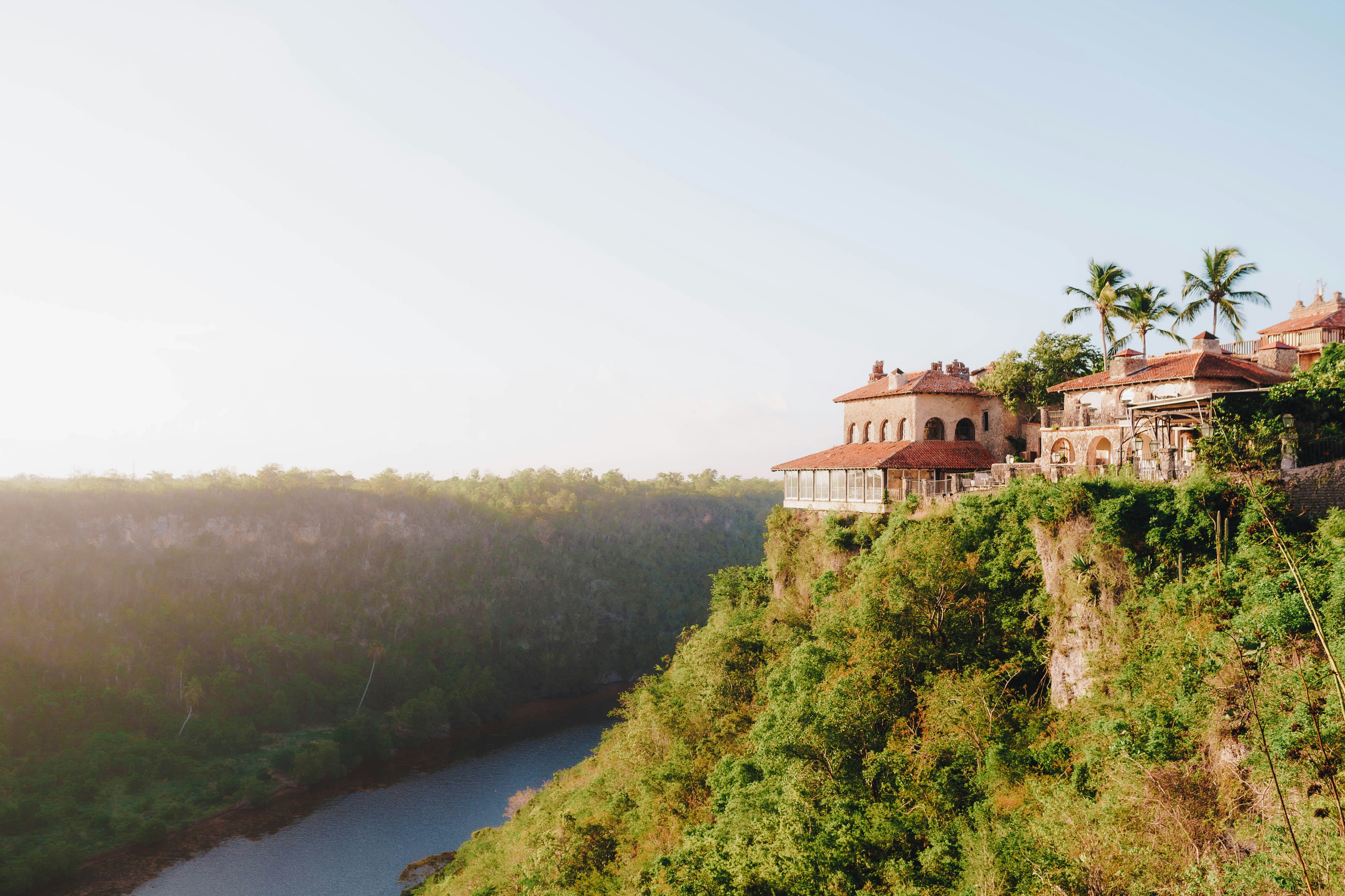 Mediterranean-style villa perched on a lush cliffside overlooking a river under a clear blue sky.