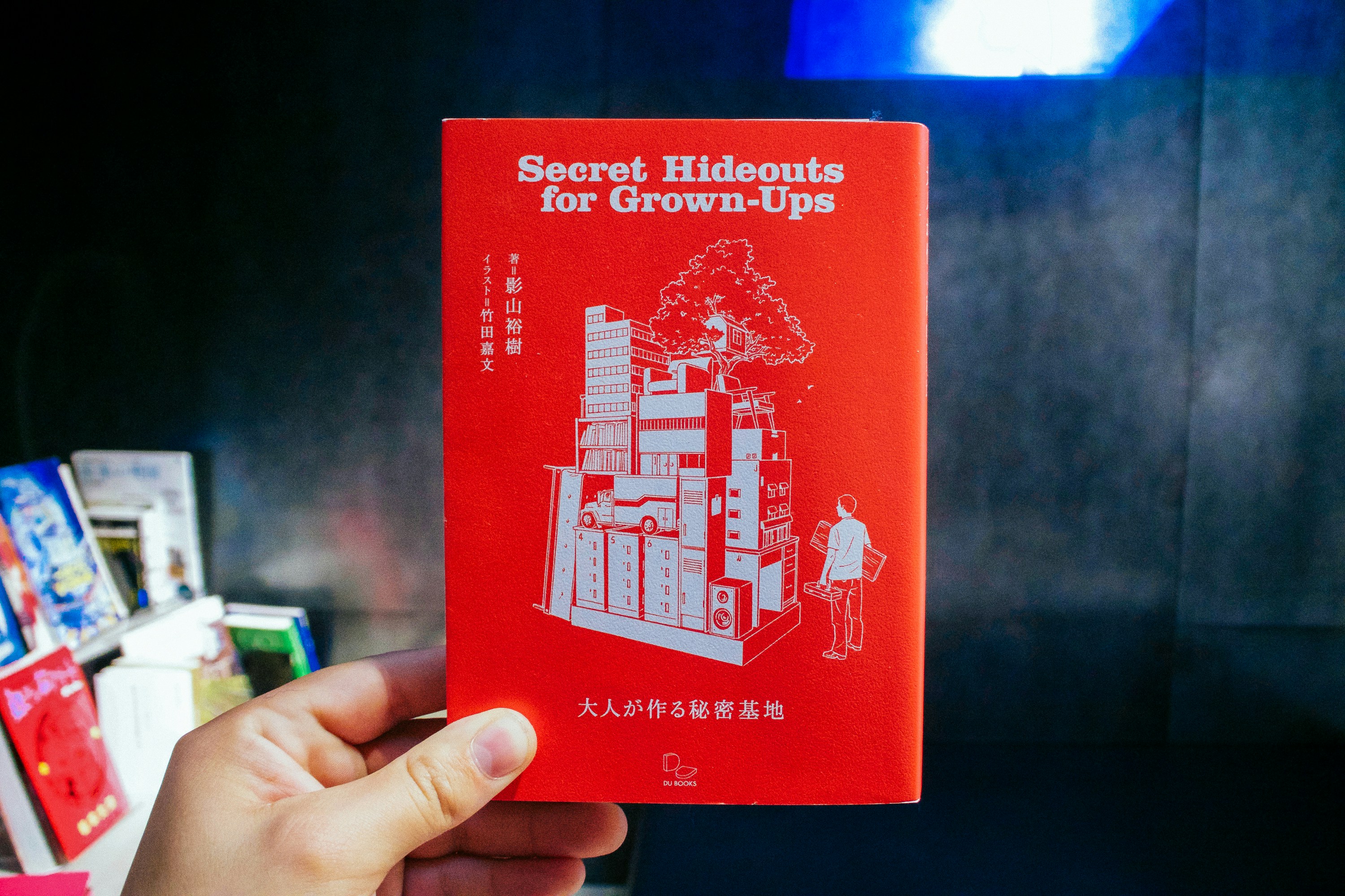 Red hardcover book cover titled 'Secret Hideouts for Grown-Ups' held by a left hand against a dark backdrop.