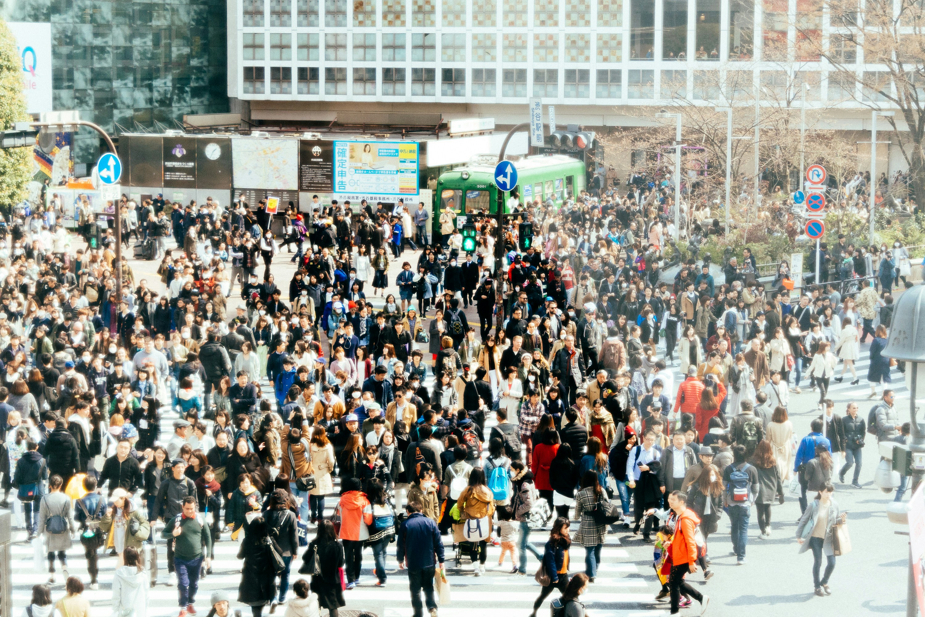 A dense crowd of pedestrians fills a busy city square in a photograph, moving in all directions beneath a modern building facade.