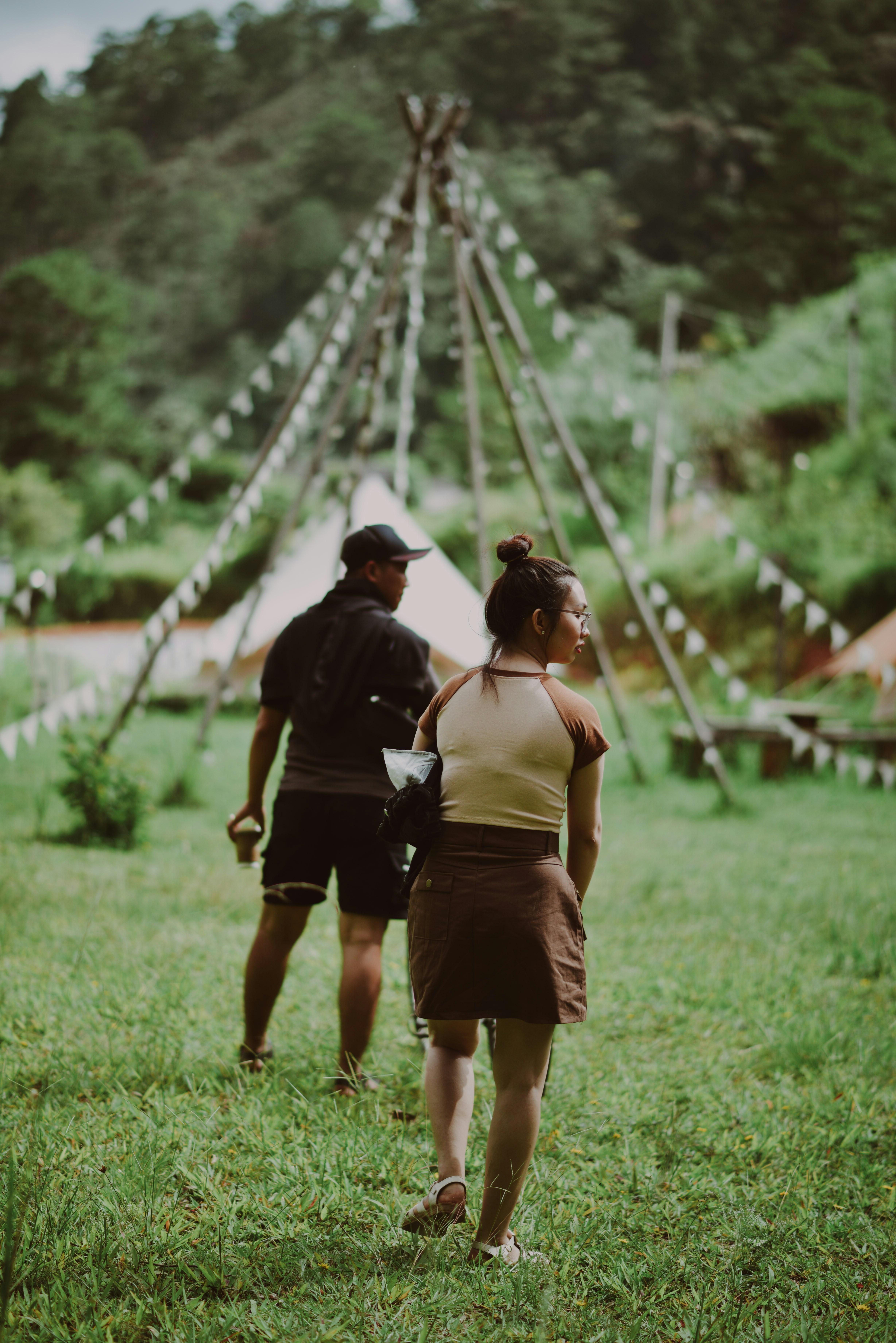 Two figures walk through a grassy festival clearing toward a central teepee framed by pennant garlands. The soft-focused photograph captures a quiet outdoor moment.