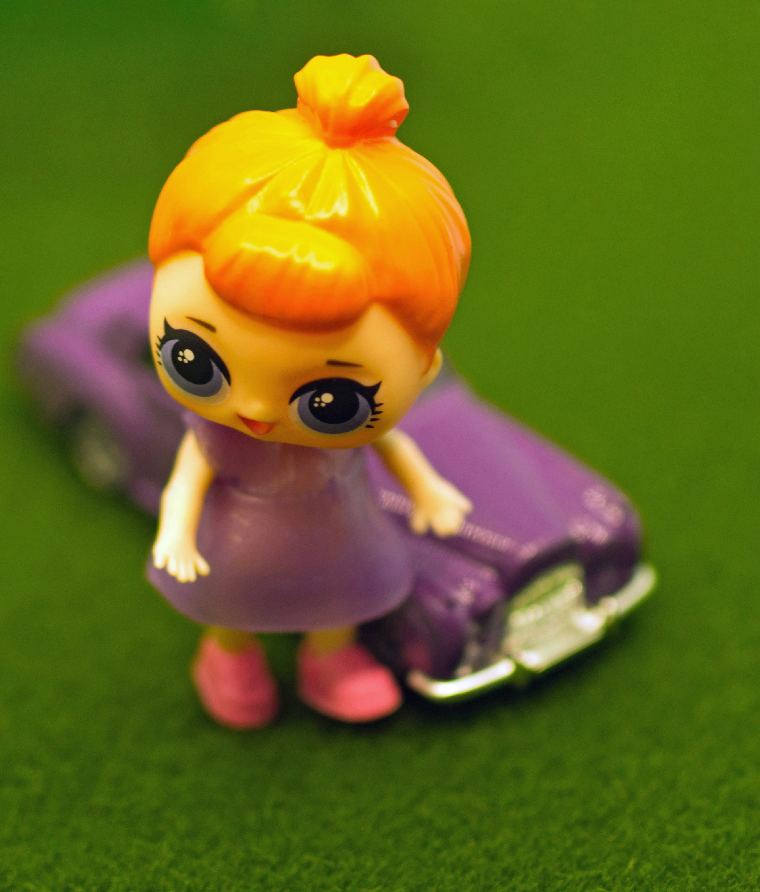 Gaze of the Tiny Orange-Haired Doll beside a Purple Car on Green Surface