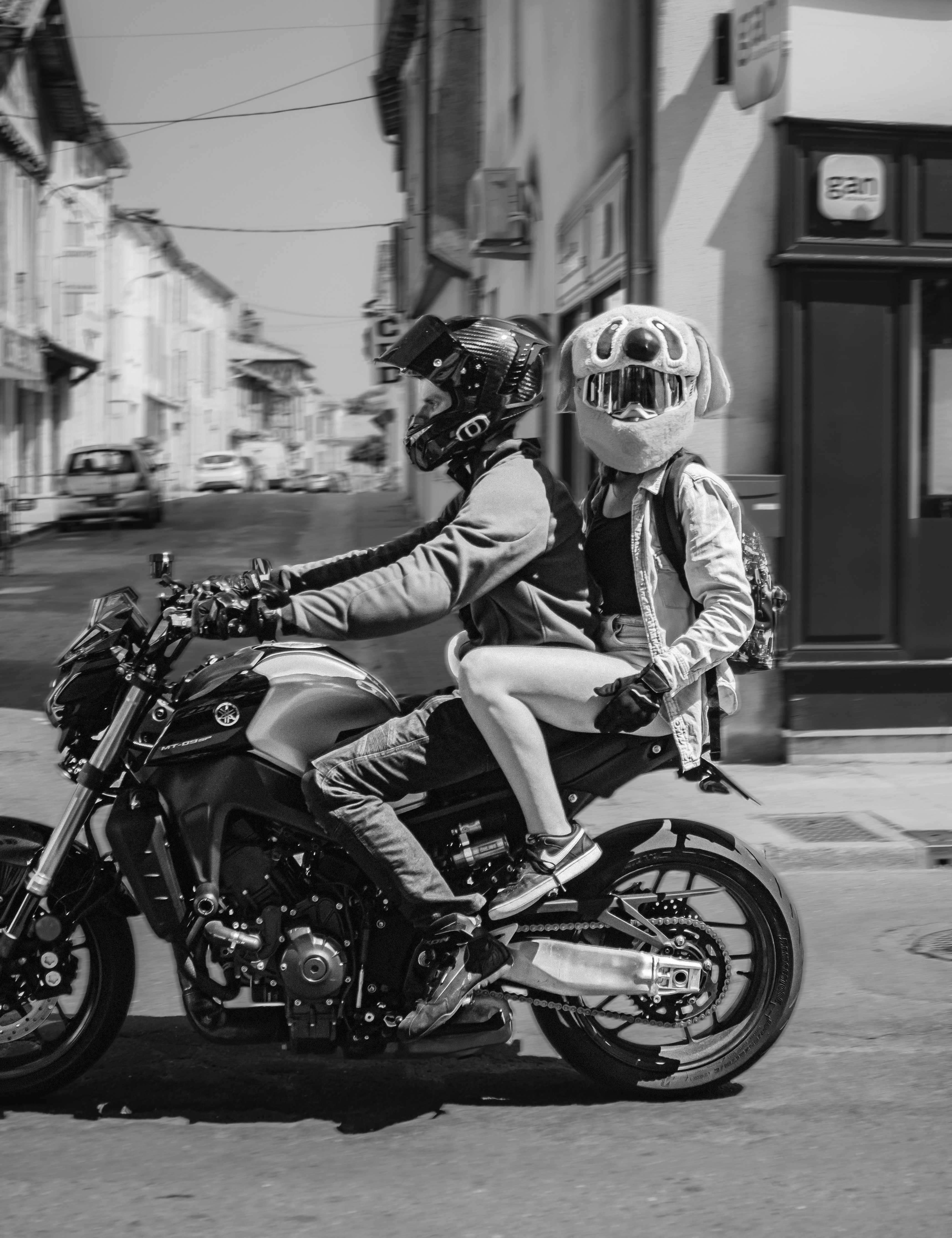 Two riders on a Yamaha motorcycle cruise a city street; the passenger wears a skull-helmet, adding a playful, eerie flair to the black-and-white shot. The image captures urban texture and motion.