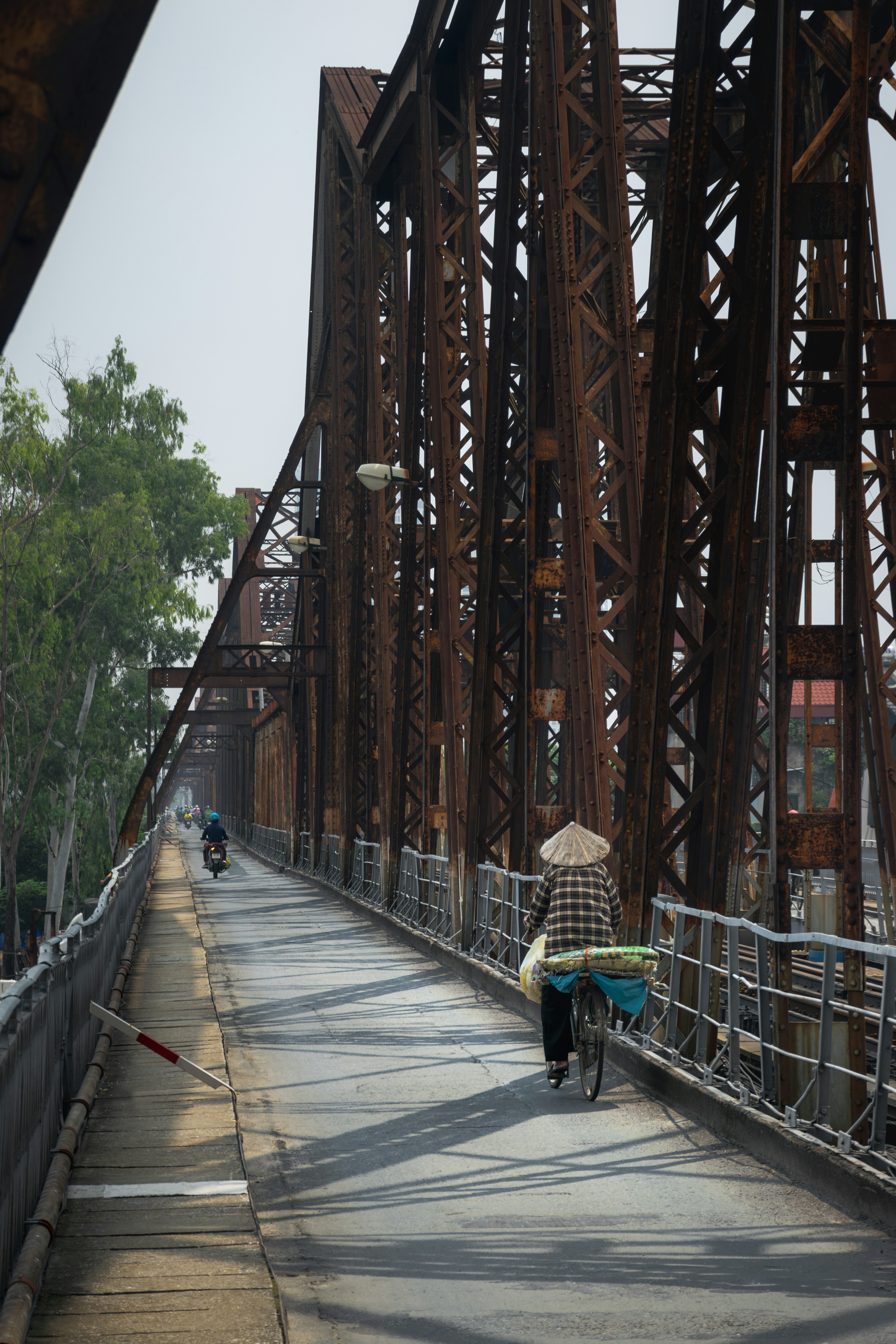 A narrow pedestrian walkway along a rusted iron truss bridge, with a conical-hatted rider pushing a bicycle in the foreground and a cyclist receding in the distance.