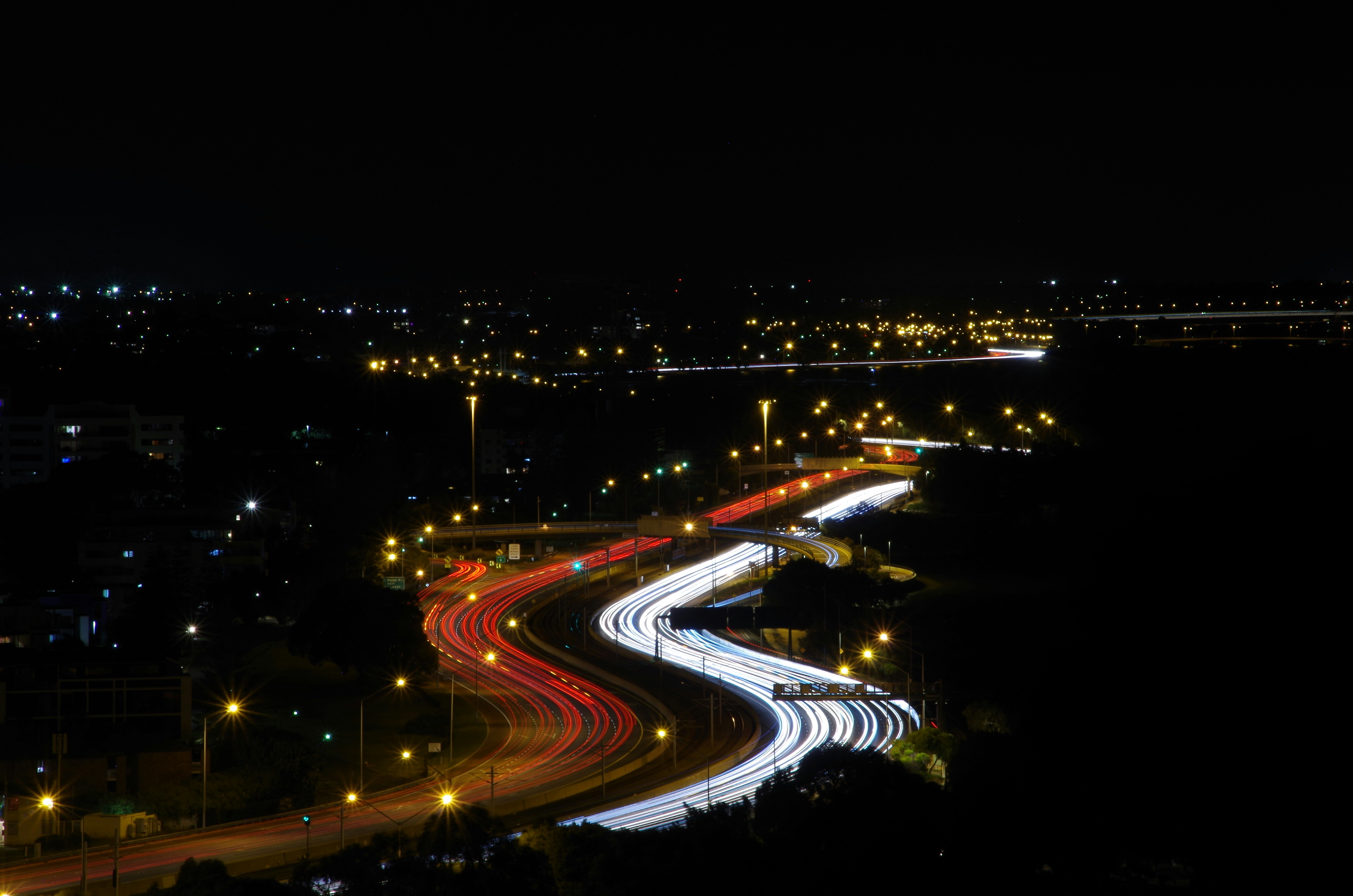 Long exposure shot capturing the flowing light trails of vehicles on a winding highway at night, contrasted against a city skyline illuminated by distant lights.