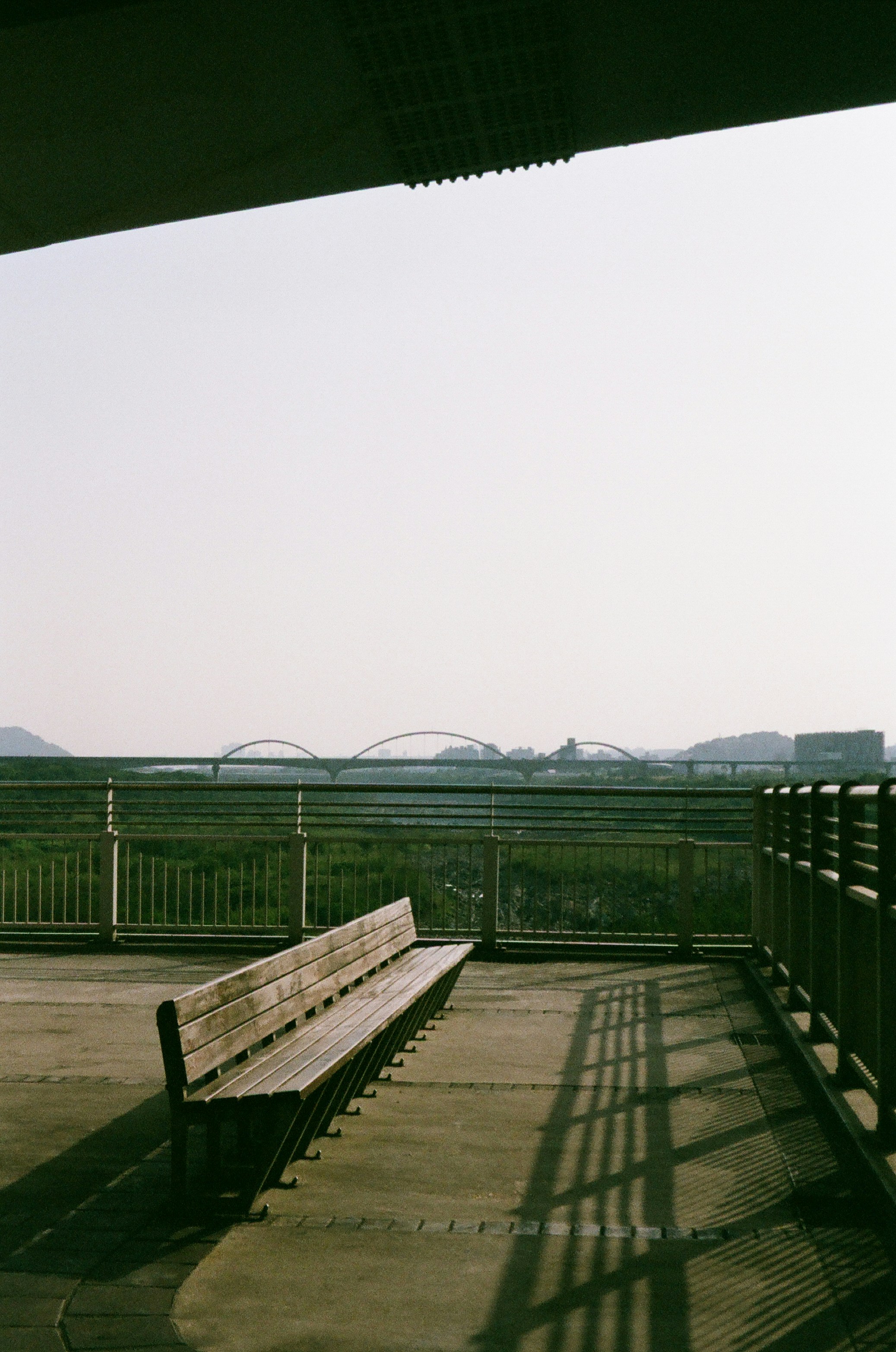 Sunlit riverside walkway with a lone bench and railing, distant curved bridge on the horizon.