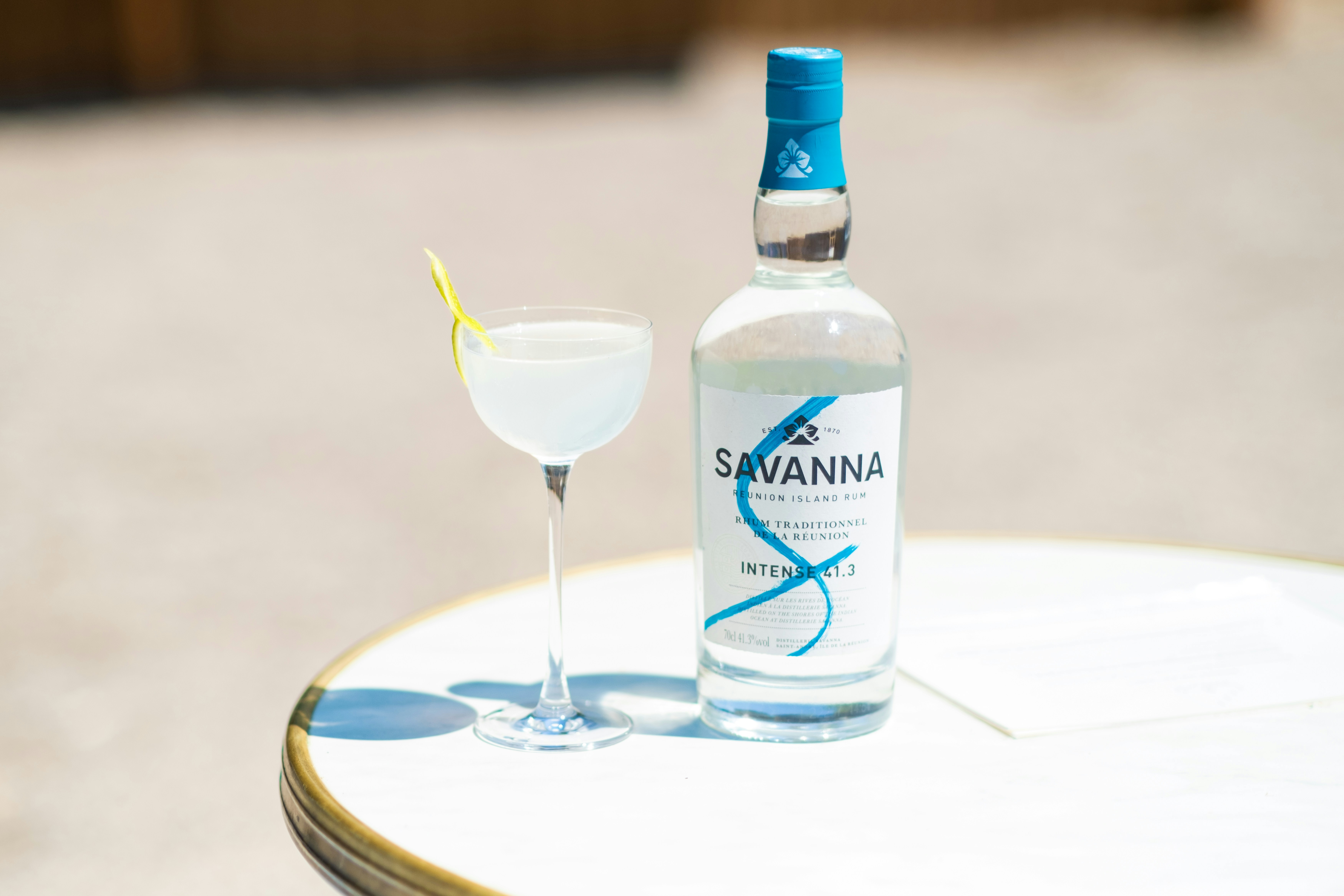 Savanna bottle with a lemon-twist cocktail on a sunlit patio table, background softly blurred.