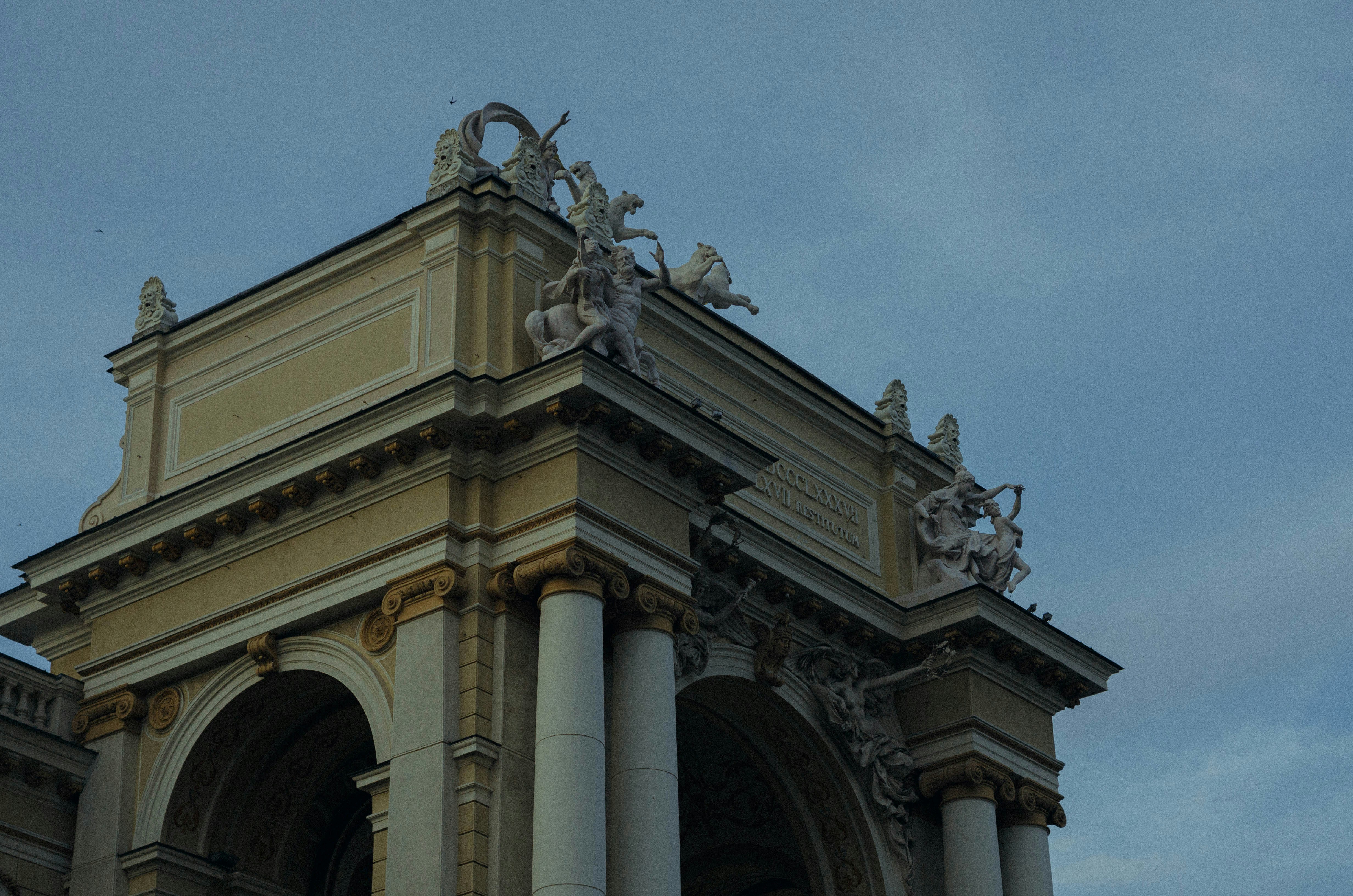 Classical building facade with ornate sculptures atop columned arches against a cloudy sky.