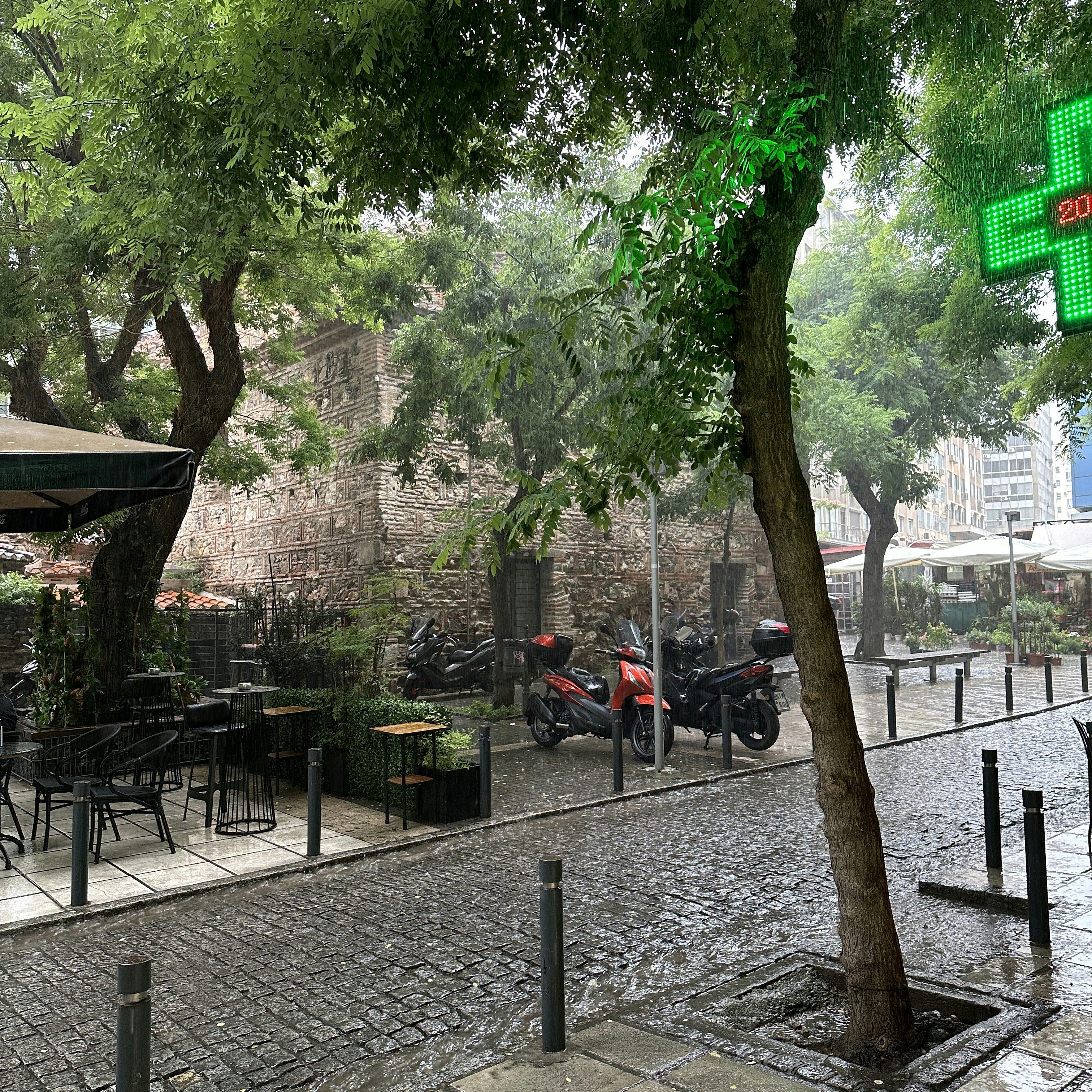 Rain-Soaked Cobblestones: Scooters Rest by a Tree-Lined Café and Stone Wall