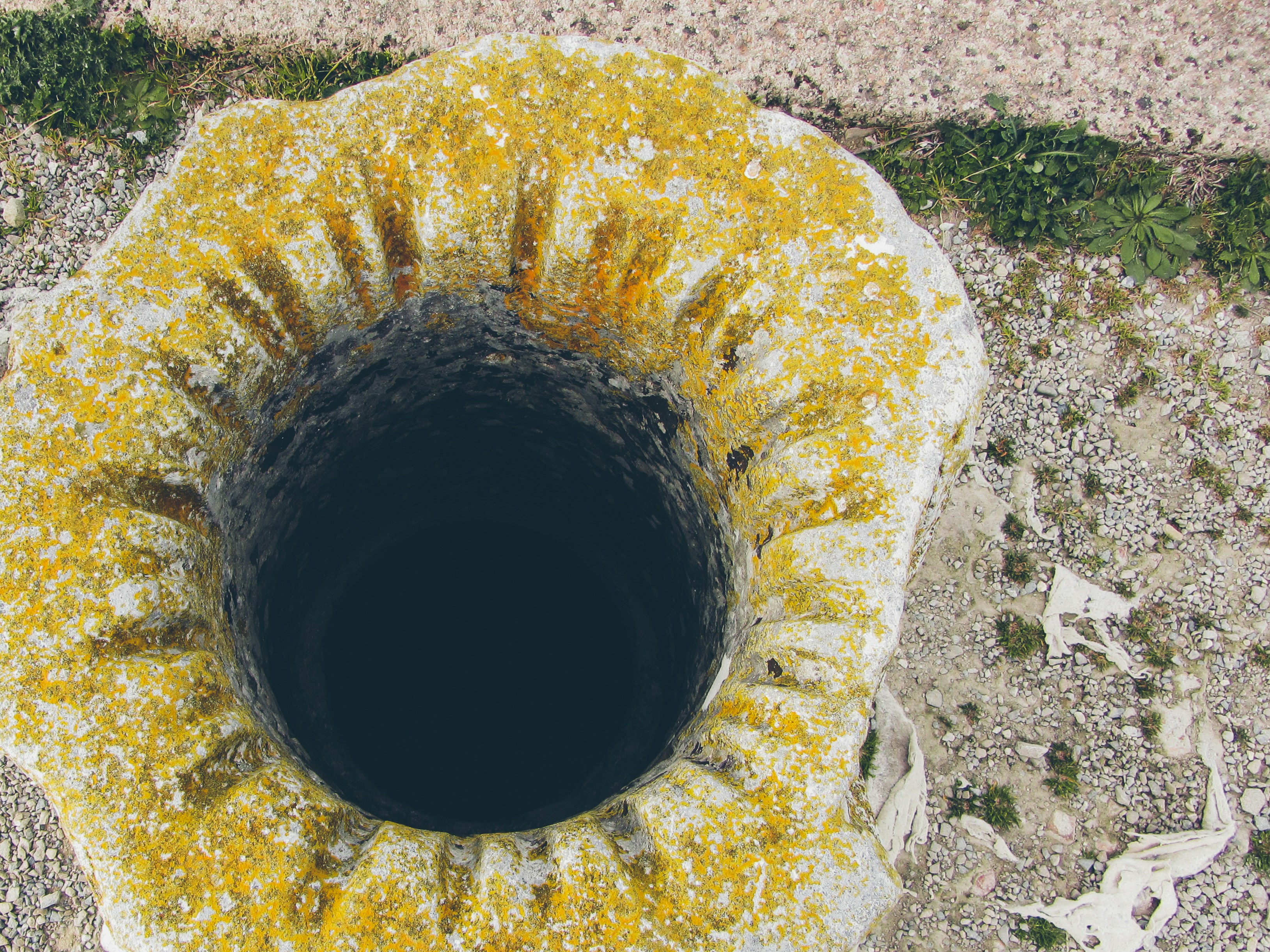 Top-down photo of a circular opening framed by a weathered stone rim and yellow moss. Rough ground surrounds the scene.