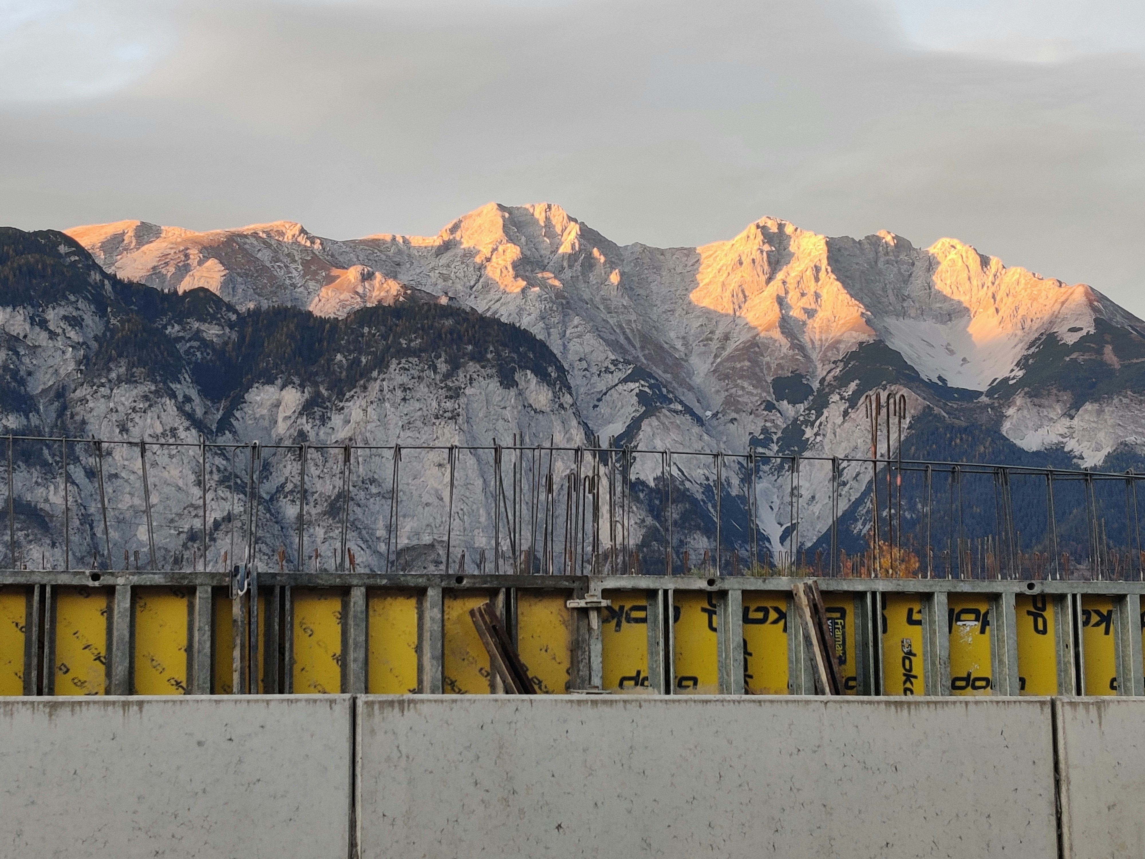 Sunlit alpine ridge rises behind a weathered construction barrier and railing, captured in a golden-hour landscape photograph.
