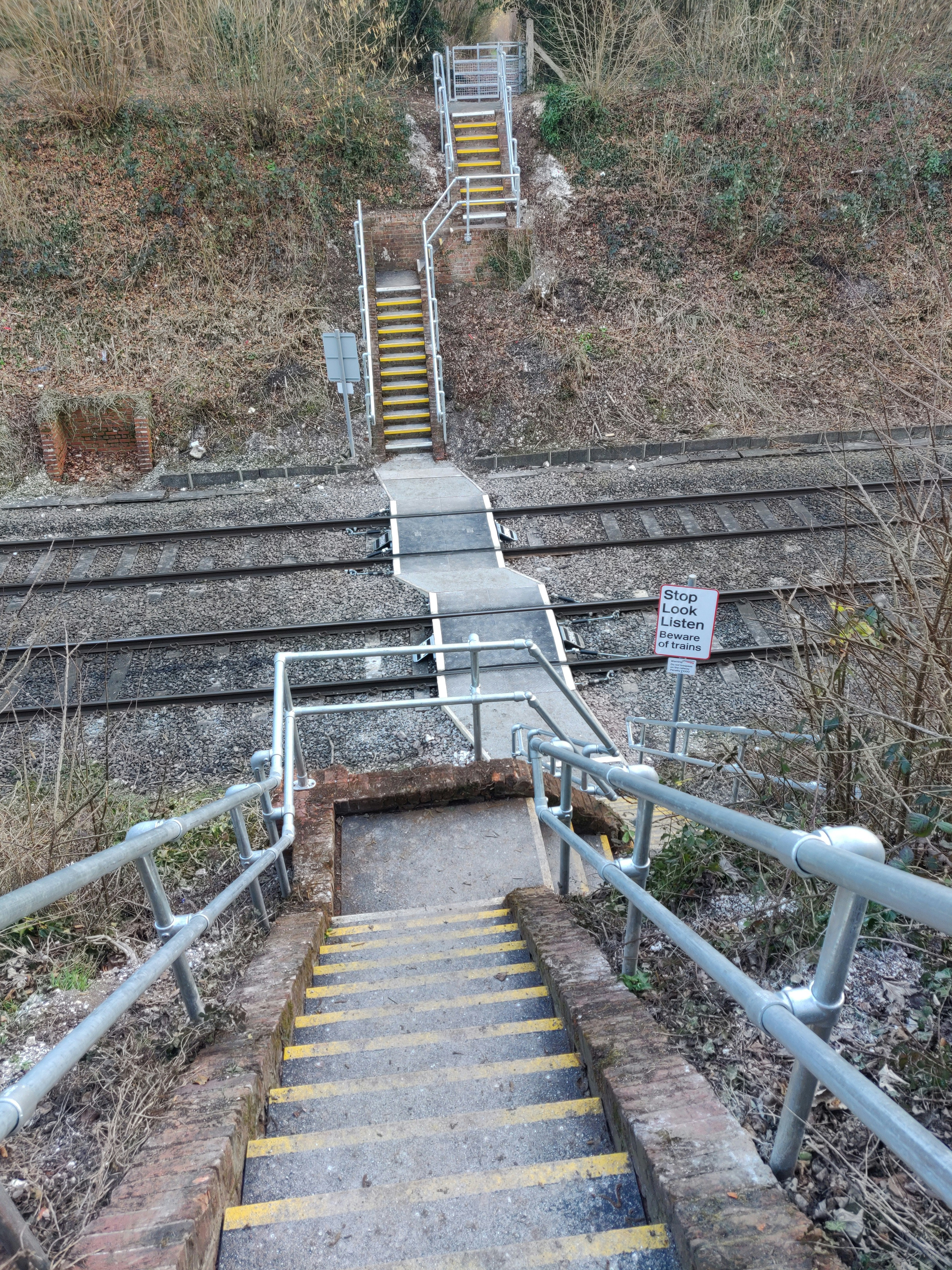 Stairs leading down to a railway crossing, flanked by a safety sign reminding pedestrians to stop, look, and listen.
