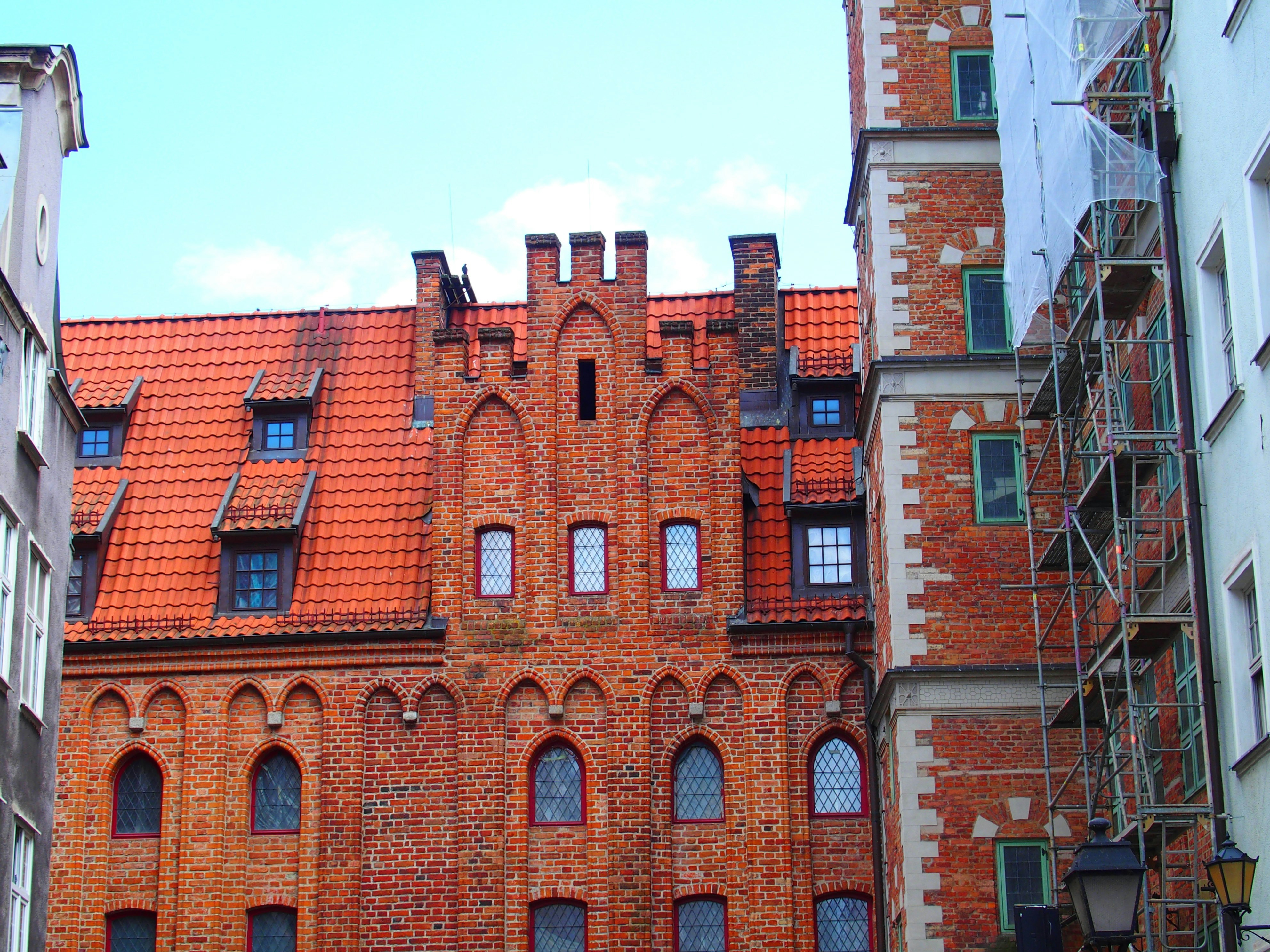 Historic brick building with distinctive Gothic architecture and vibrant orange roof, framed by modern structures. Renovation work is evident on the adjacent building.