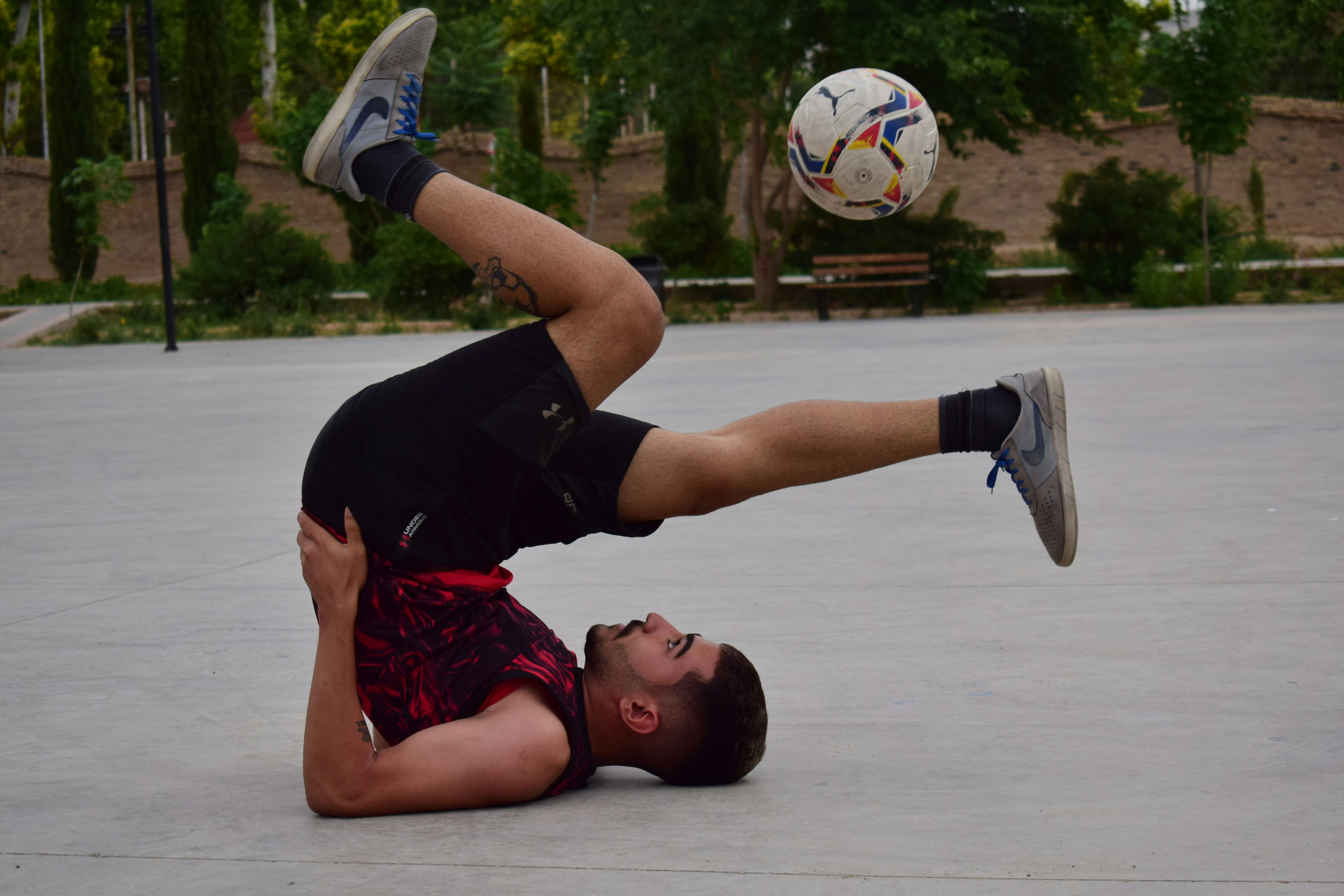 Athlete executing an acrobatic soccer trick on a concrete court with trees in the background.