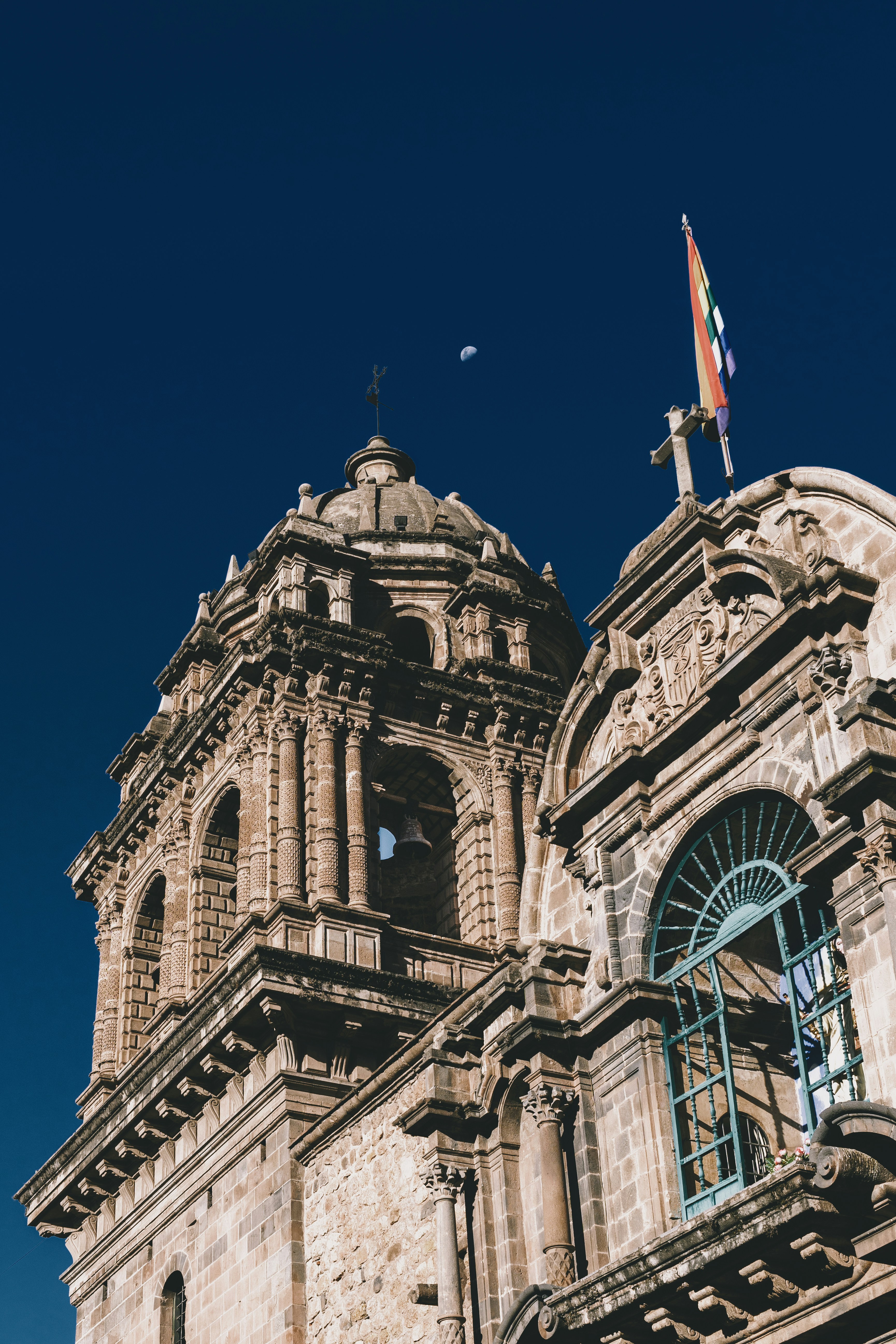 Historic church tower adorned with a vibrant flag against a deep blue sky, featuring intricate stonework and a glimpse of the moon above.